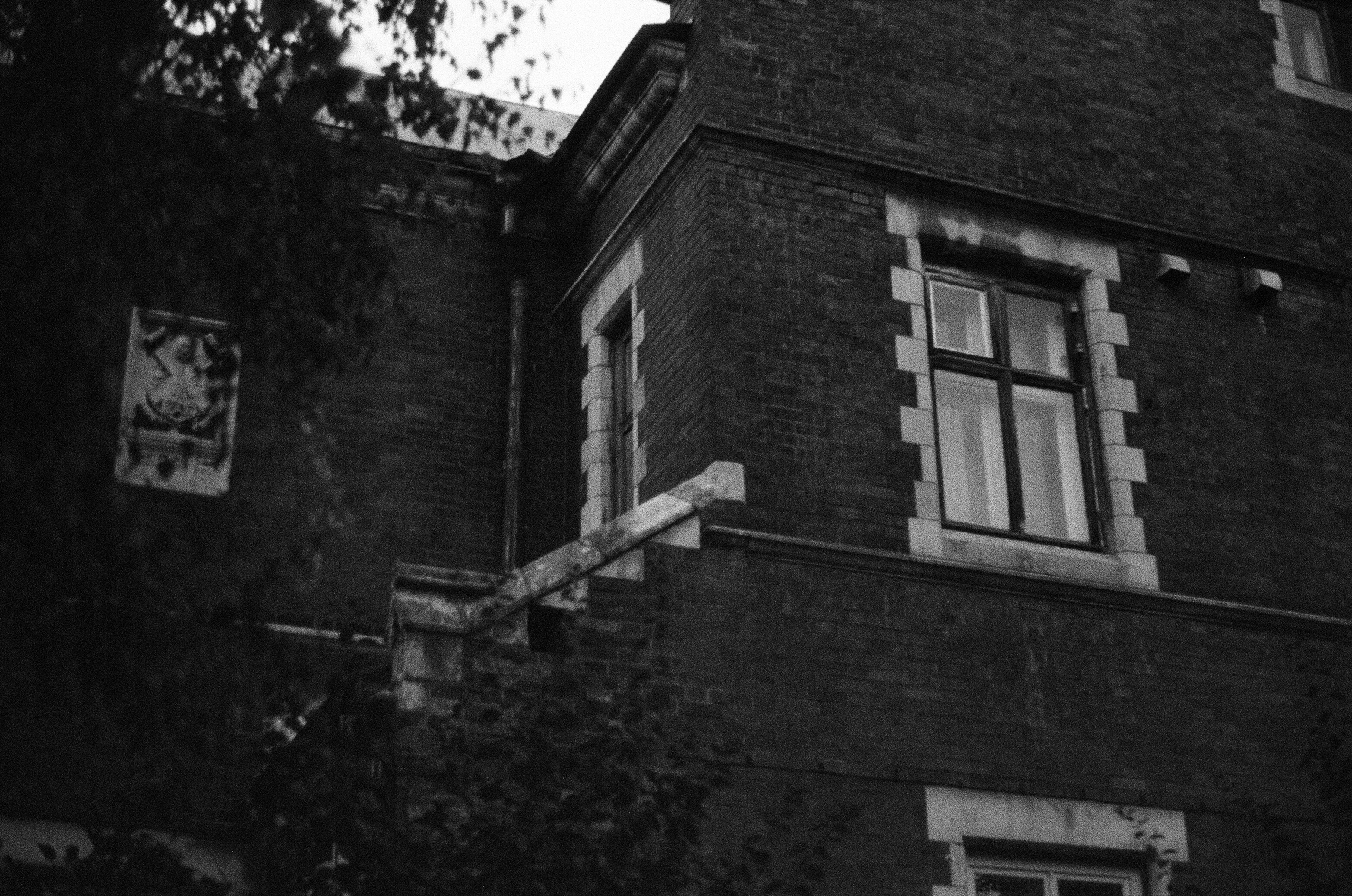This moody black‑and‑white photograph captures a corner of a weathered brick building, highlighting its austere urban character. Textured masonry, stone‑framed windows, and a diagonal wooden brace add structural tension, while overgrown foliage and a faded plaque introduce layers of decay and history.