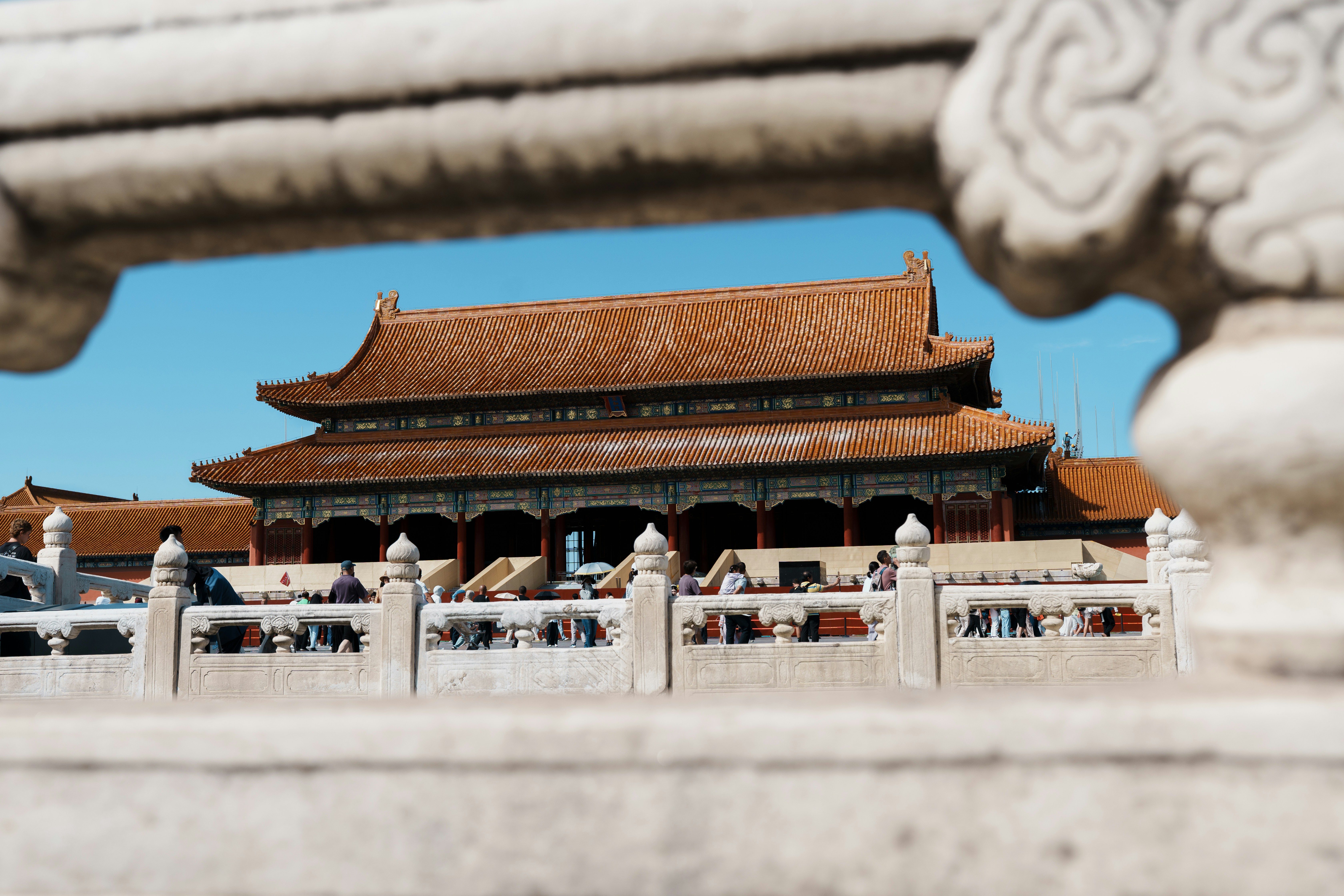 A traditional Chinese palace hall sits beyond a carved stone railing, framed by ornate stonework in the foreground; a bright blue sky and crowds are visible in the courtyard.