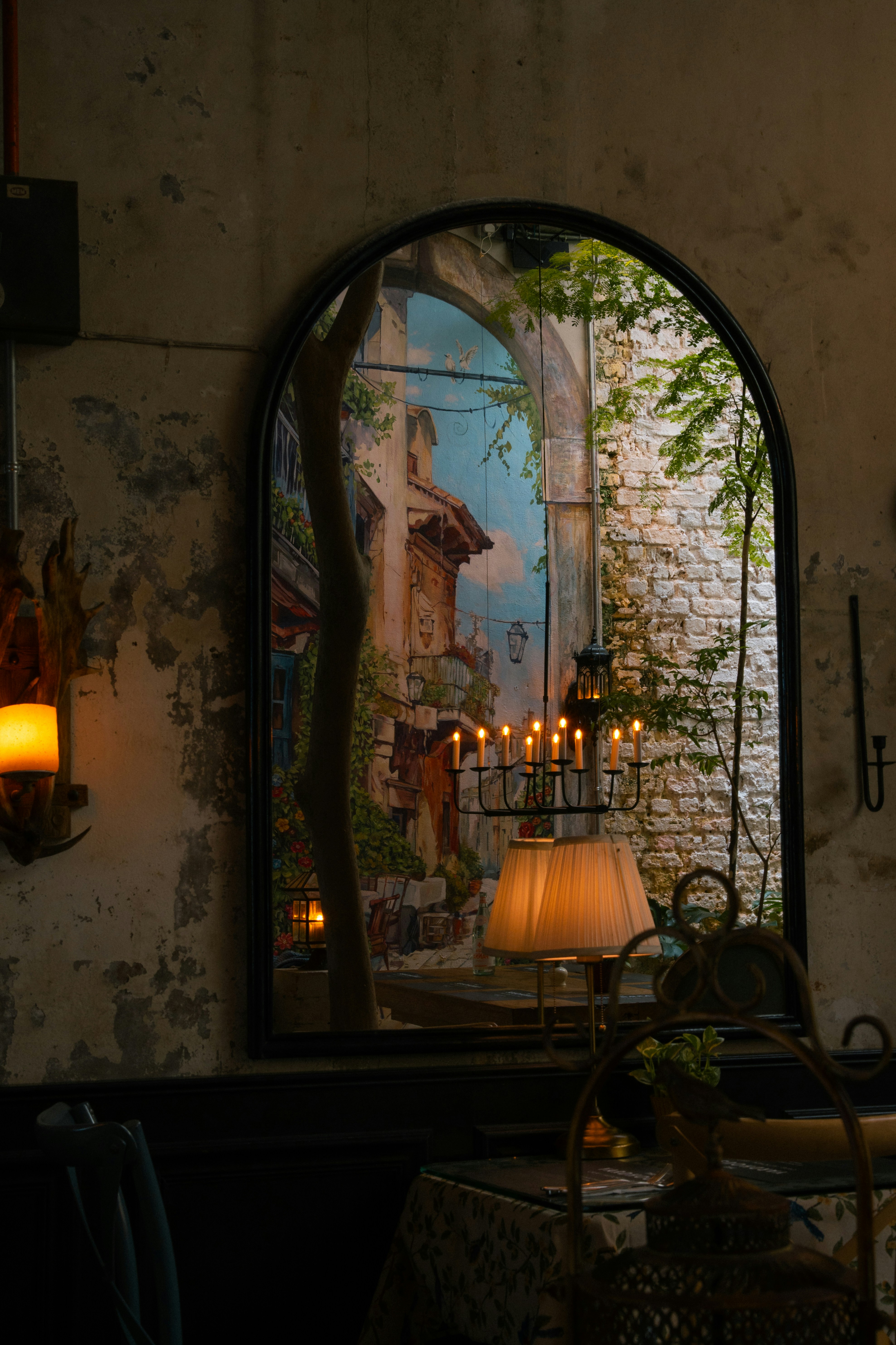 Warm interior cafe scene reflected in a tall arched window, candlelit chandelier and ambient lamp glow; a textured wall and outdoor mural visible through the arch.
