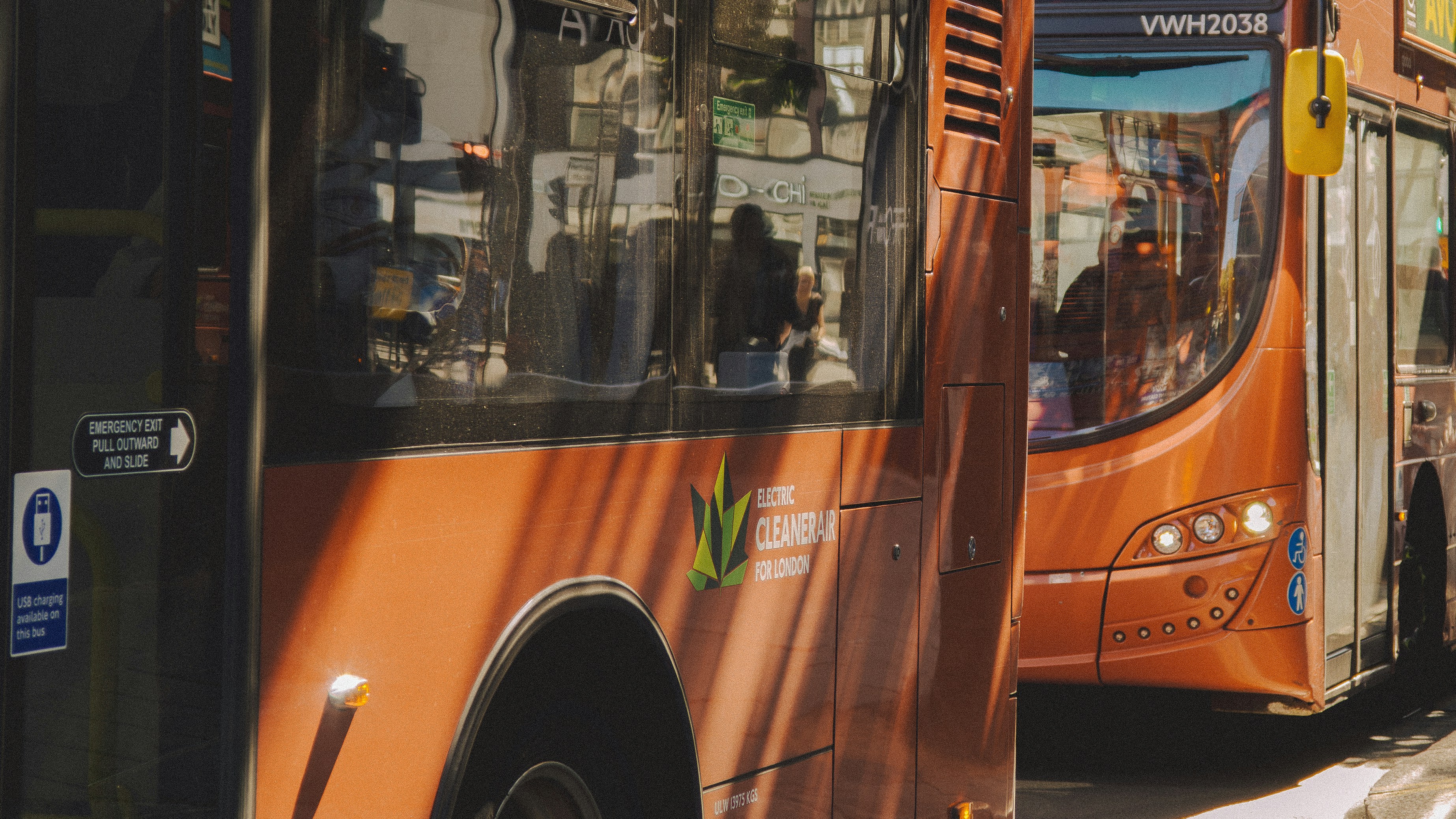 Two orange city buses overlap at a stop, with reflections and passengers visible through the windows, conveying a busy urban moment.