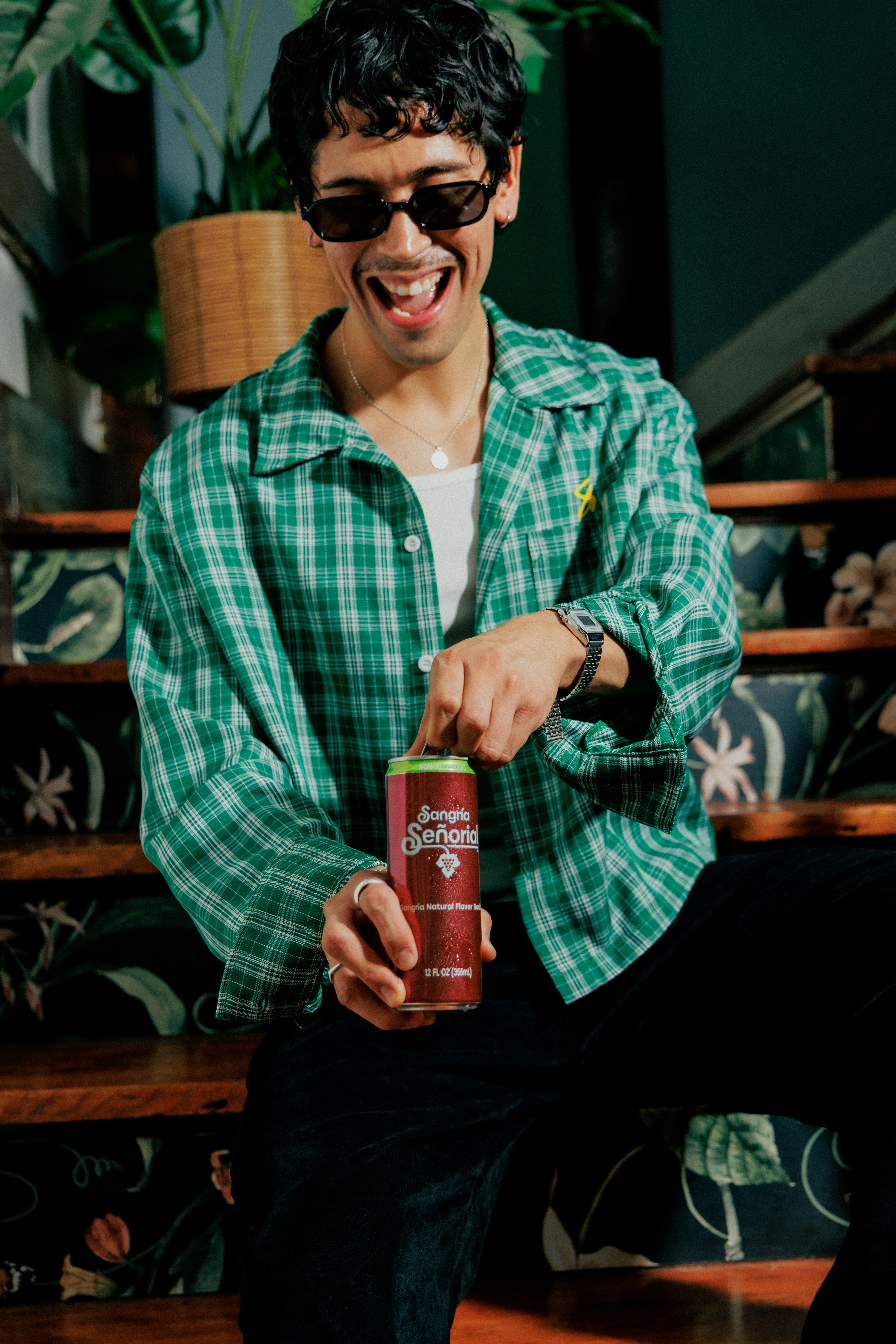 Energetic portrait of a man in a green plaid shirt, sitting on wooden stairs and opening a red soda can; the scene shows a playful, casual indoor setting with leafy decor.