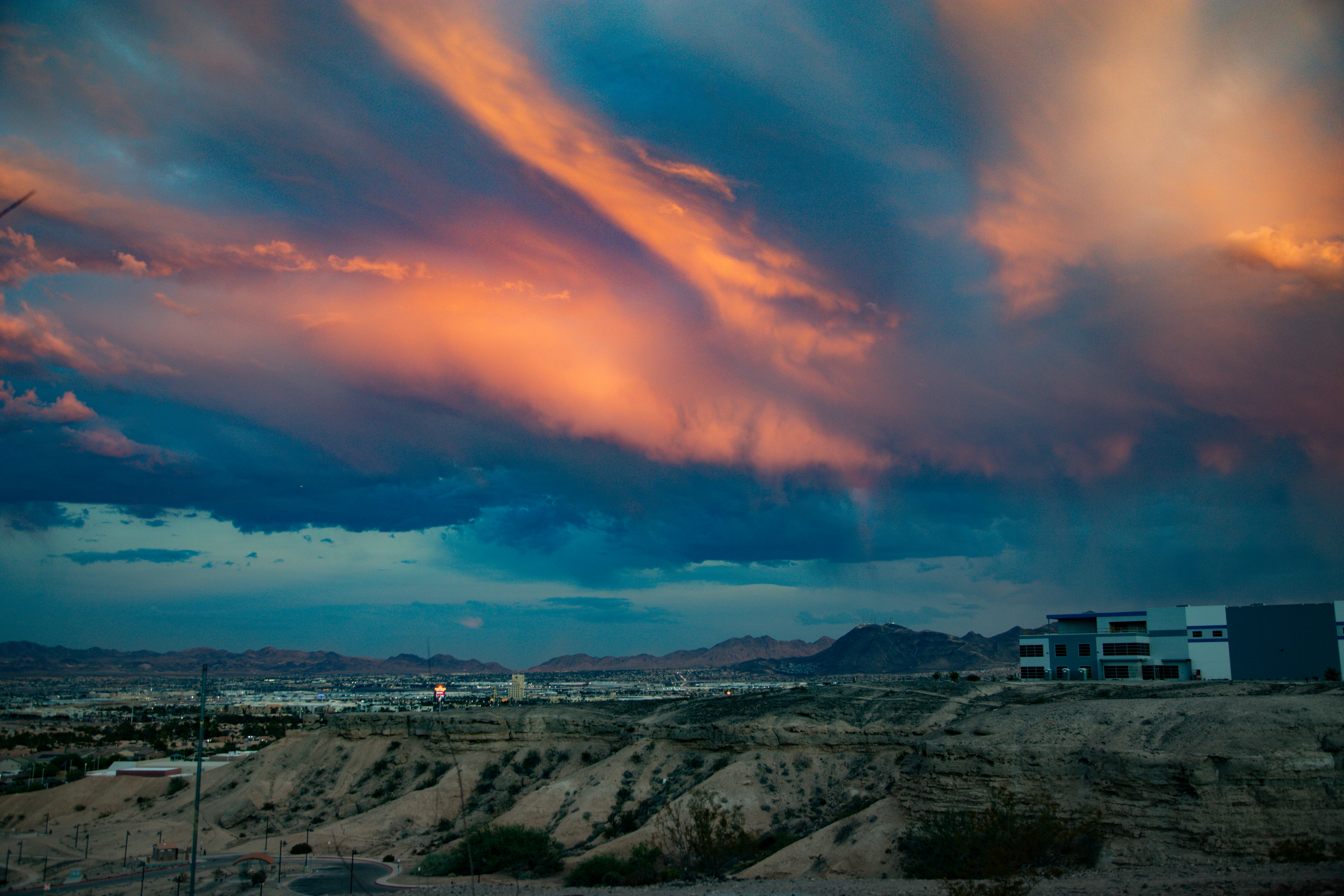 Roaring sunset clouds drift over a desert hillside with distant buildings, capturing a quiet urban fringe at twilight.
