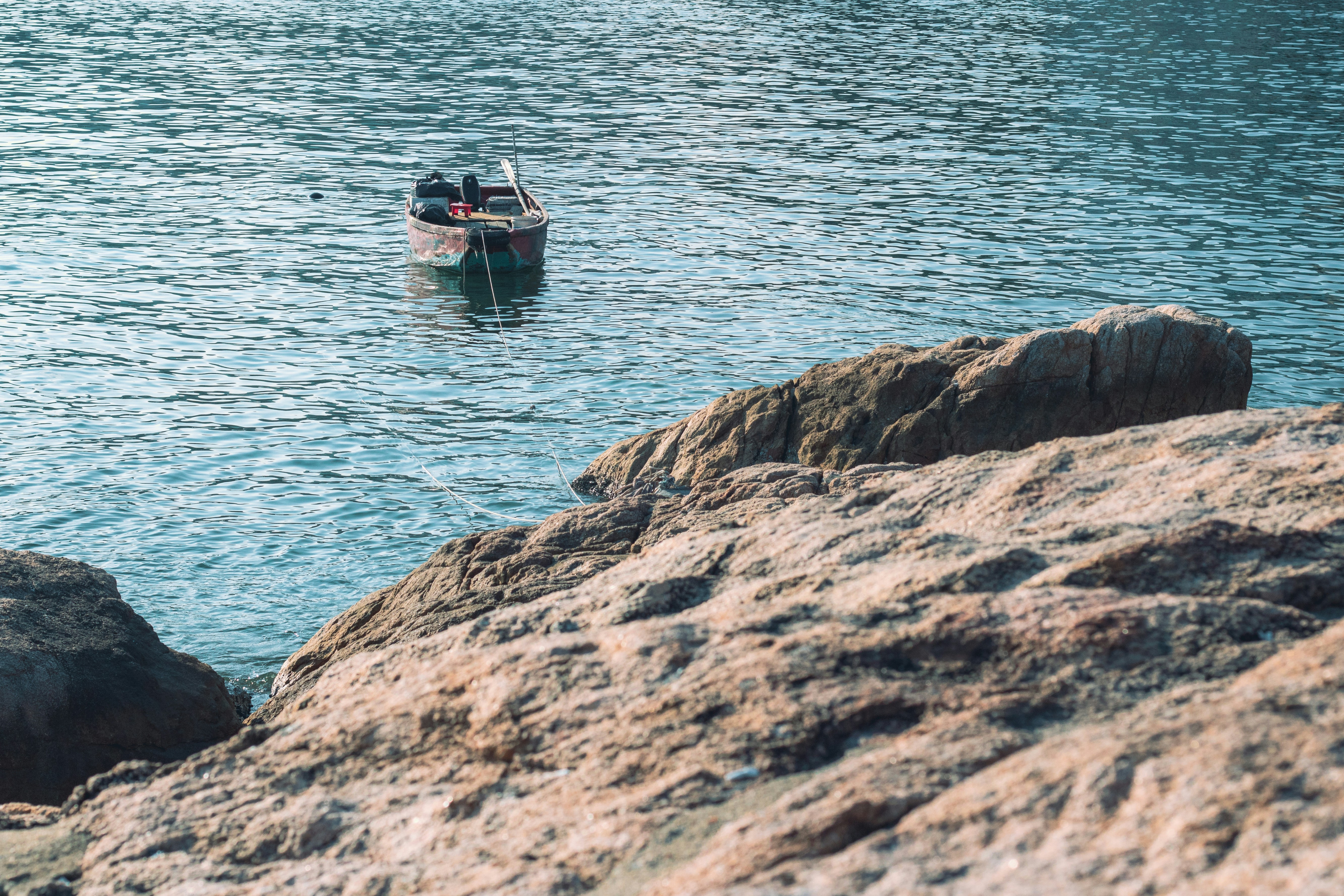 Small boat rests near rocky coastline with turquoise water in the background, highlighting the contrast between smooth water and textured rock formations. A minimal seascape with a solitary craft.
