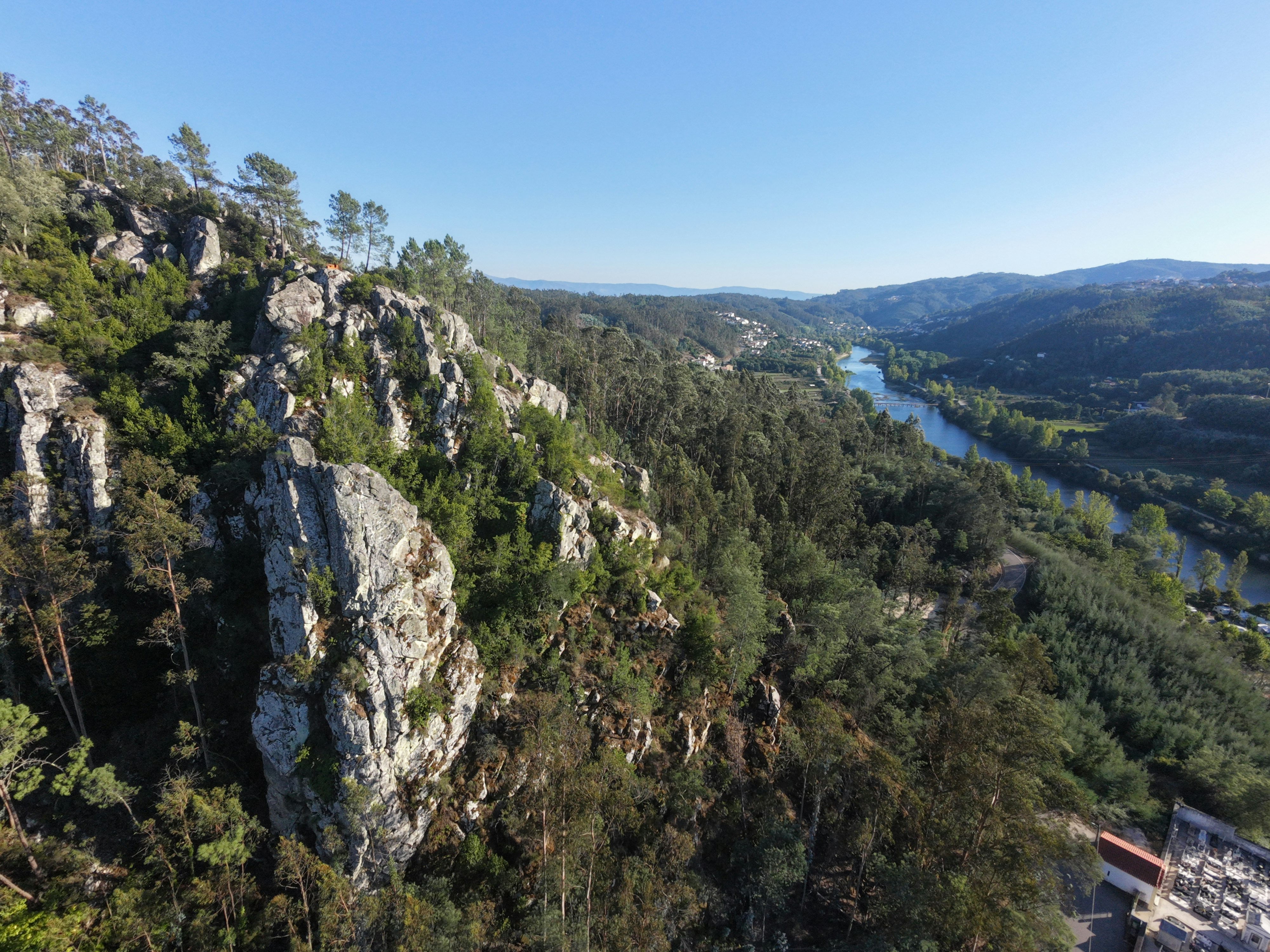 Cliffside pines overlook a winding river through a valley, with rugged rock faces and distant hills under a clear sky. A riverside landscape captured from a high vantage point.