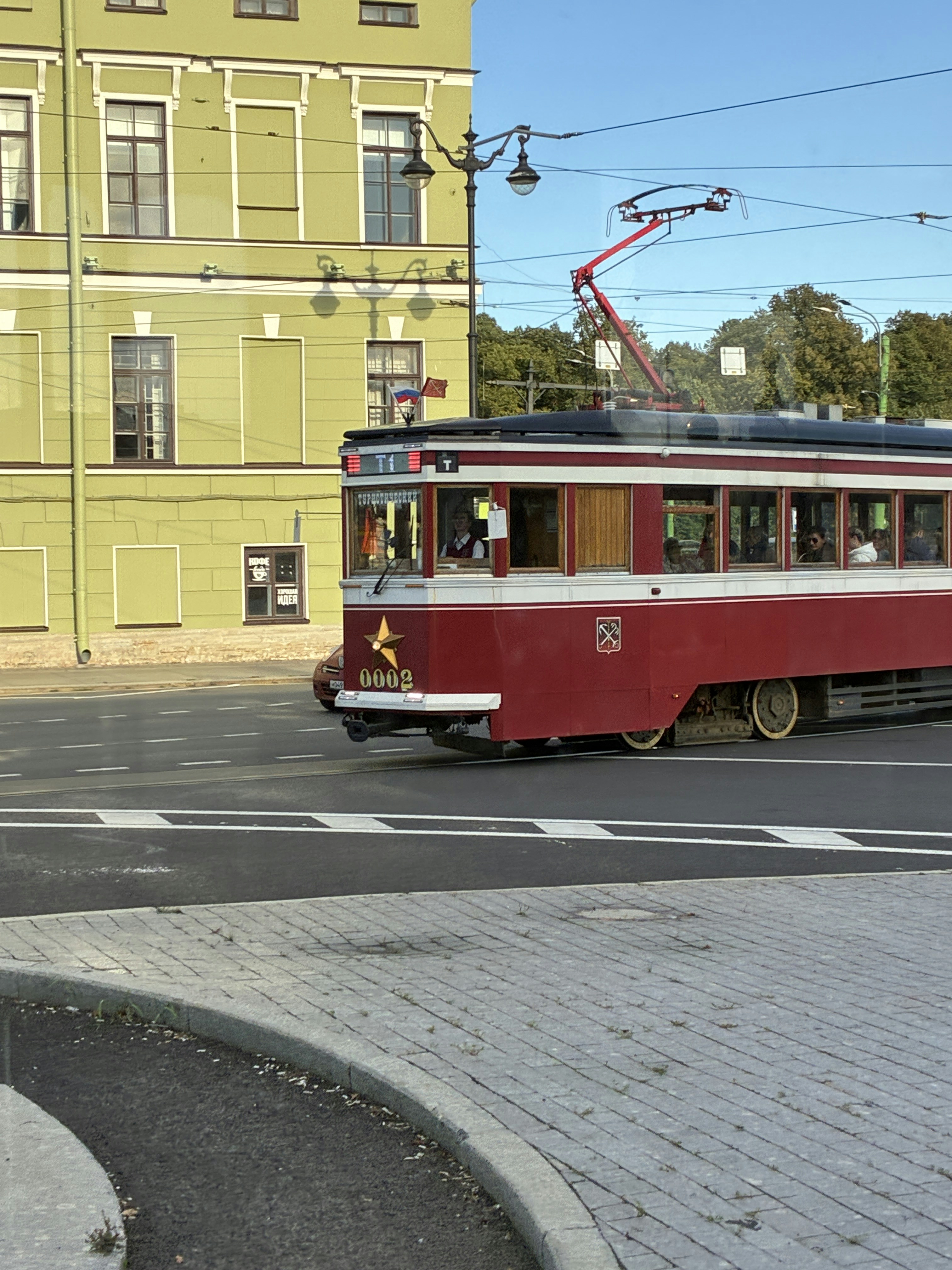 Red tram crossing a sunlit street beside a yellow-green building; foreground shows curved pavement and cobblestones, passengers visible through windows.