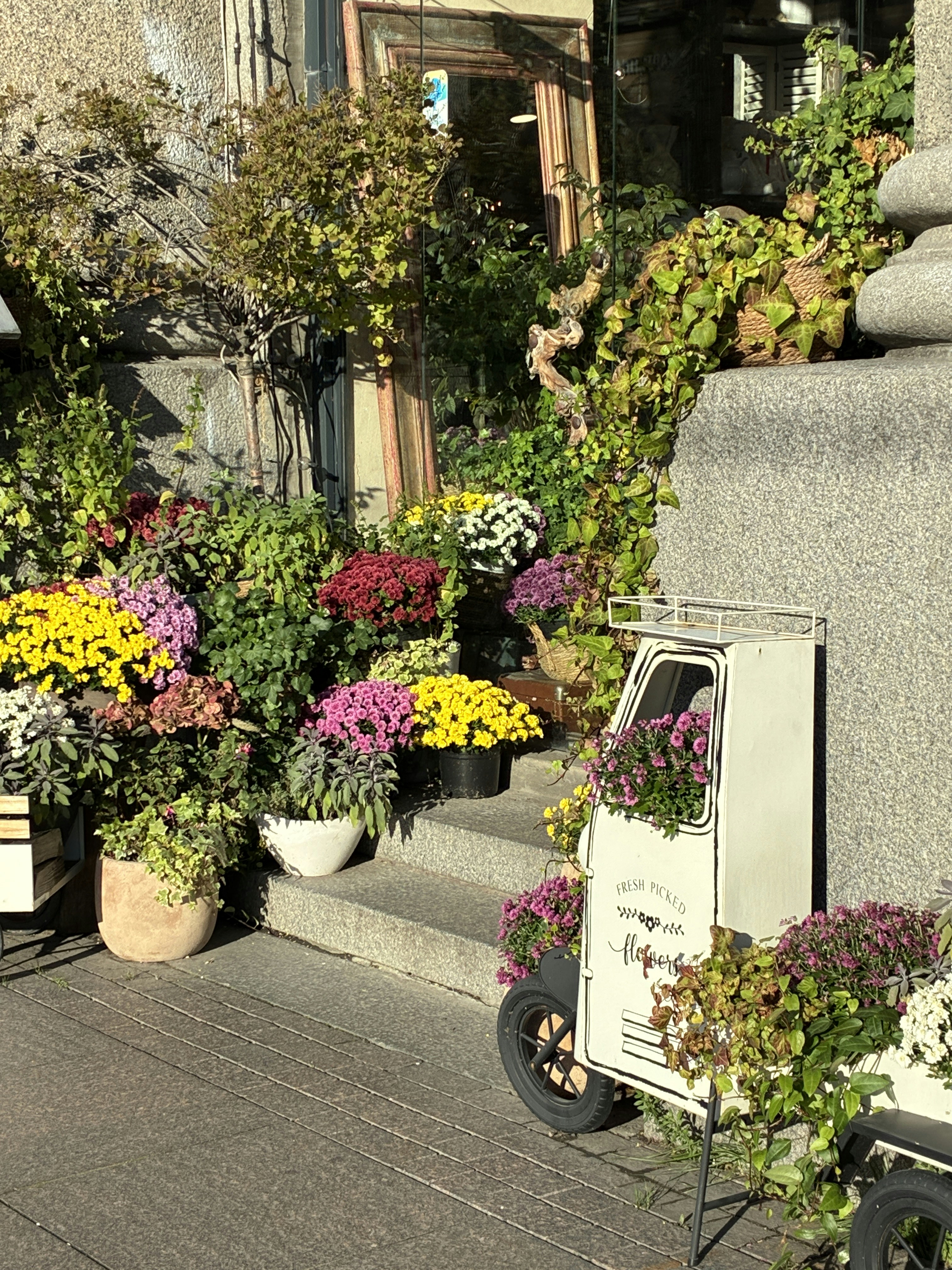 Colorful potted flowers line a stone staircase beside a white vintage cart, creating a charming floral display.