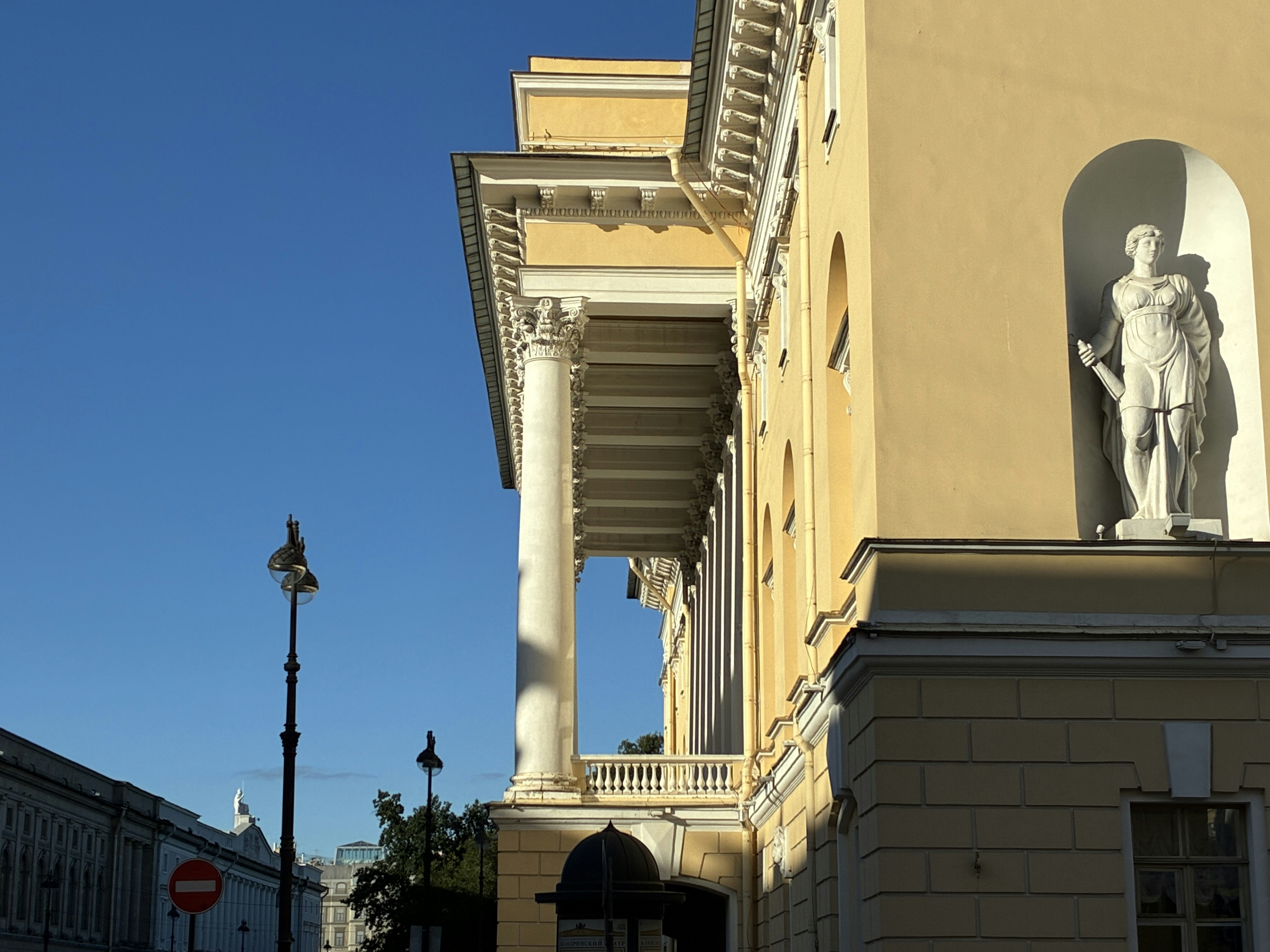 Sunlit yellow neoclassical building with arches and a statue in an alcove; street scene with a lamppost and blue sky.