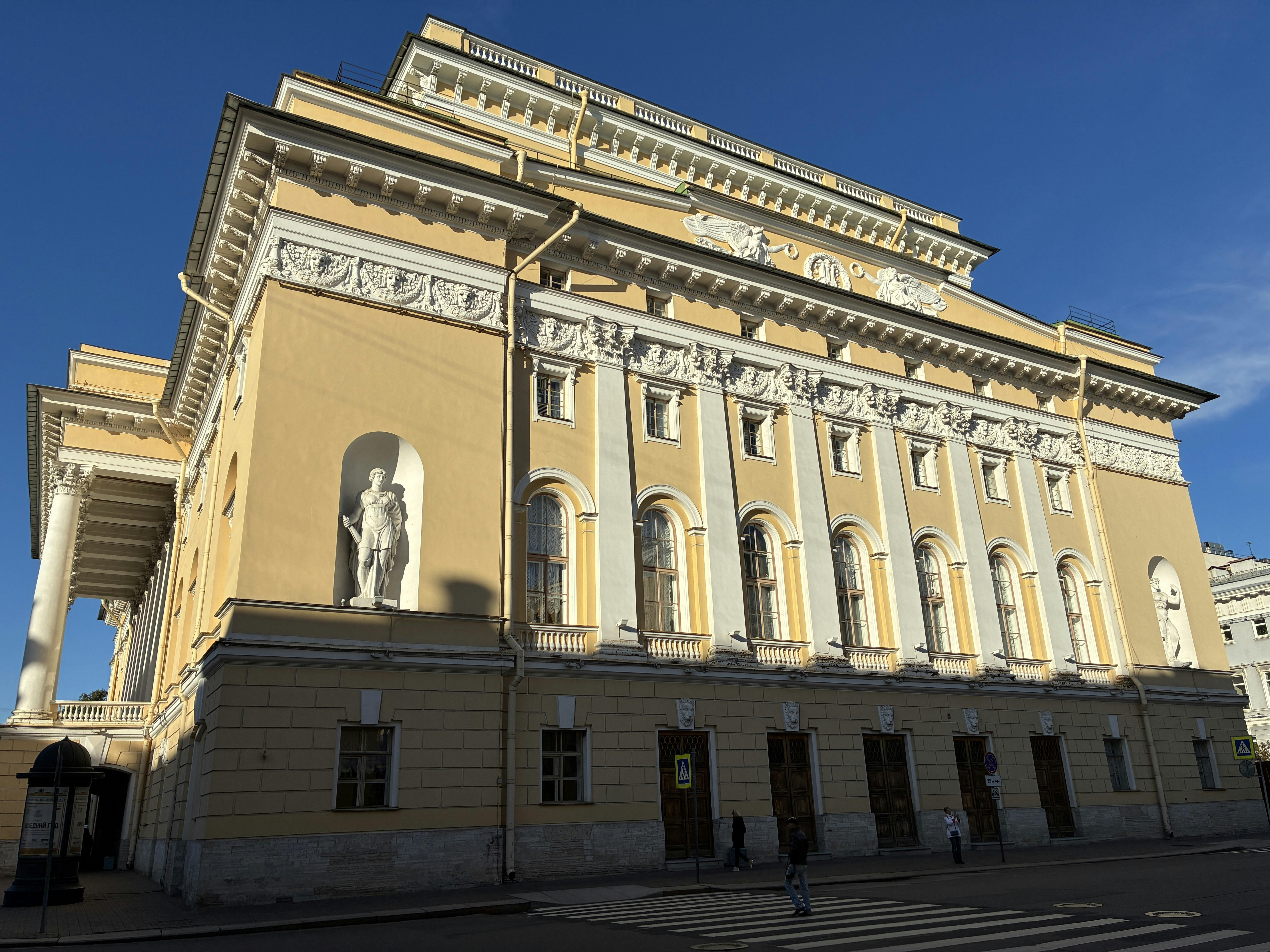 Sunlit neoclassical building with ornate friezes and statues along a broad street; a historic civic facade bathed in afternoon light.