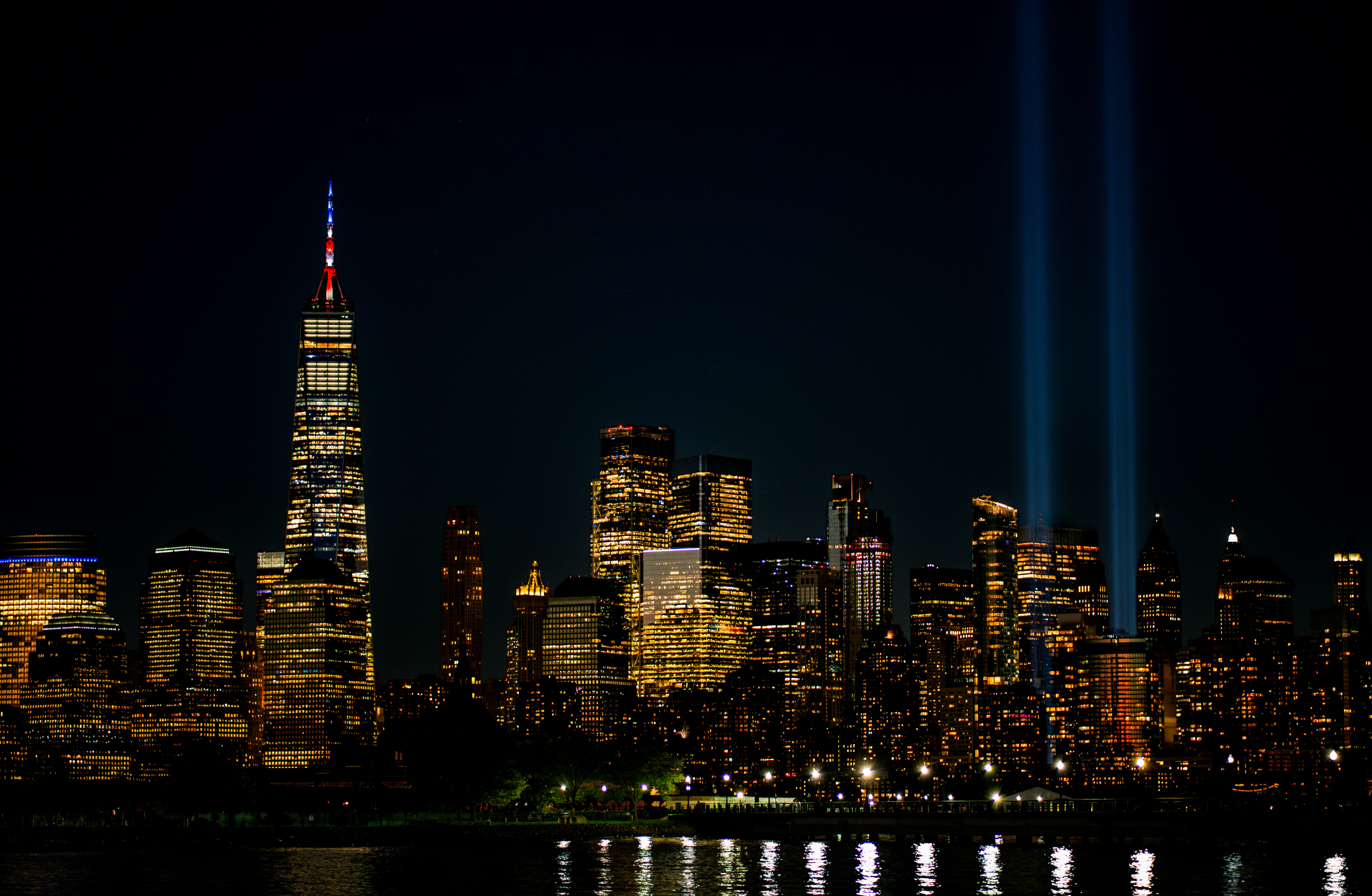 Night skyline of Manhattan with illuminated skyscrapers reflecting on dark water; a calm cityscape capturing the glow of high-rises and the Twin Towers memorial beams in the distance. This is a cityscape photograph.