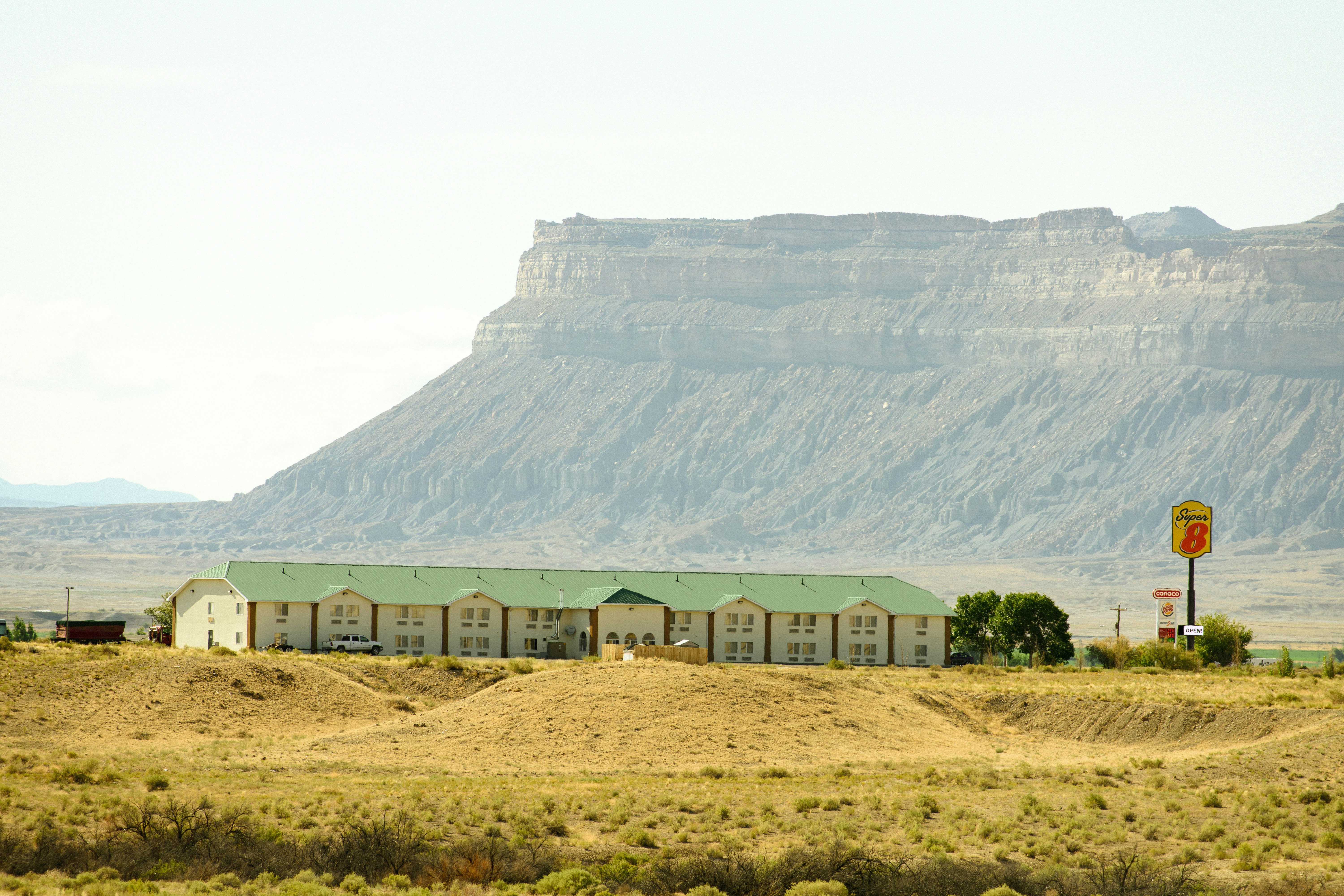 Low horizon view of a single-row of light-colored houses with green roofs in arid terrain, backed by a layered plateau; a small roadside sign hints at a rural stop. (Log in the scene resembles a housing complex in desert landscape.)