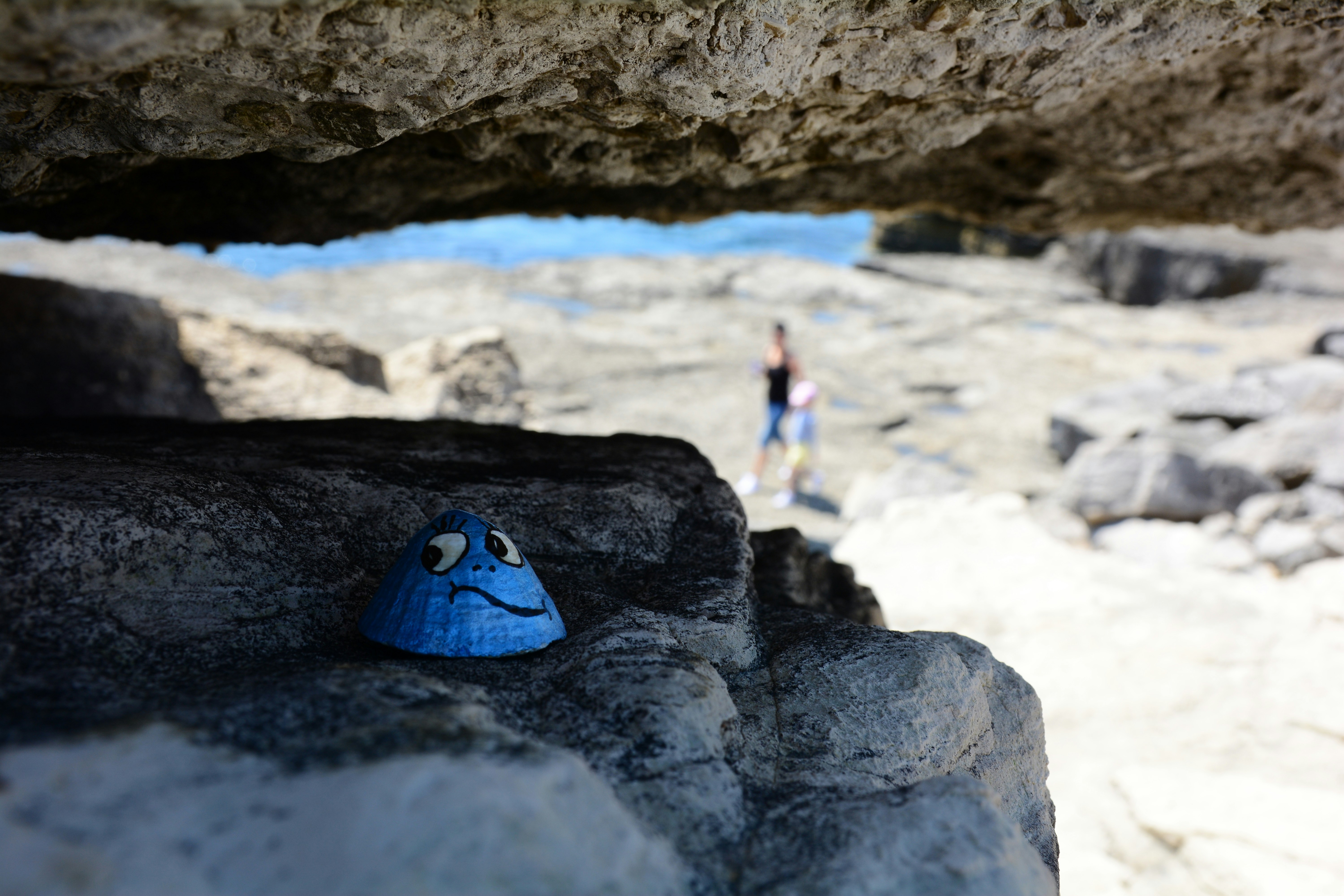 A small blue smiley rock sits in a dark crevice at a rocky shoreline, with a distant blurred figure on the sunlit beach beyond. The scene emphasizes contrast between shaded foreground and bright background.
