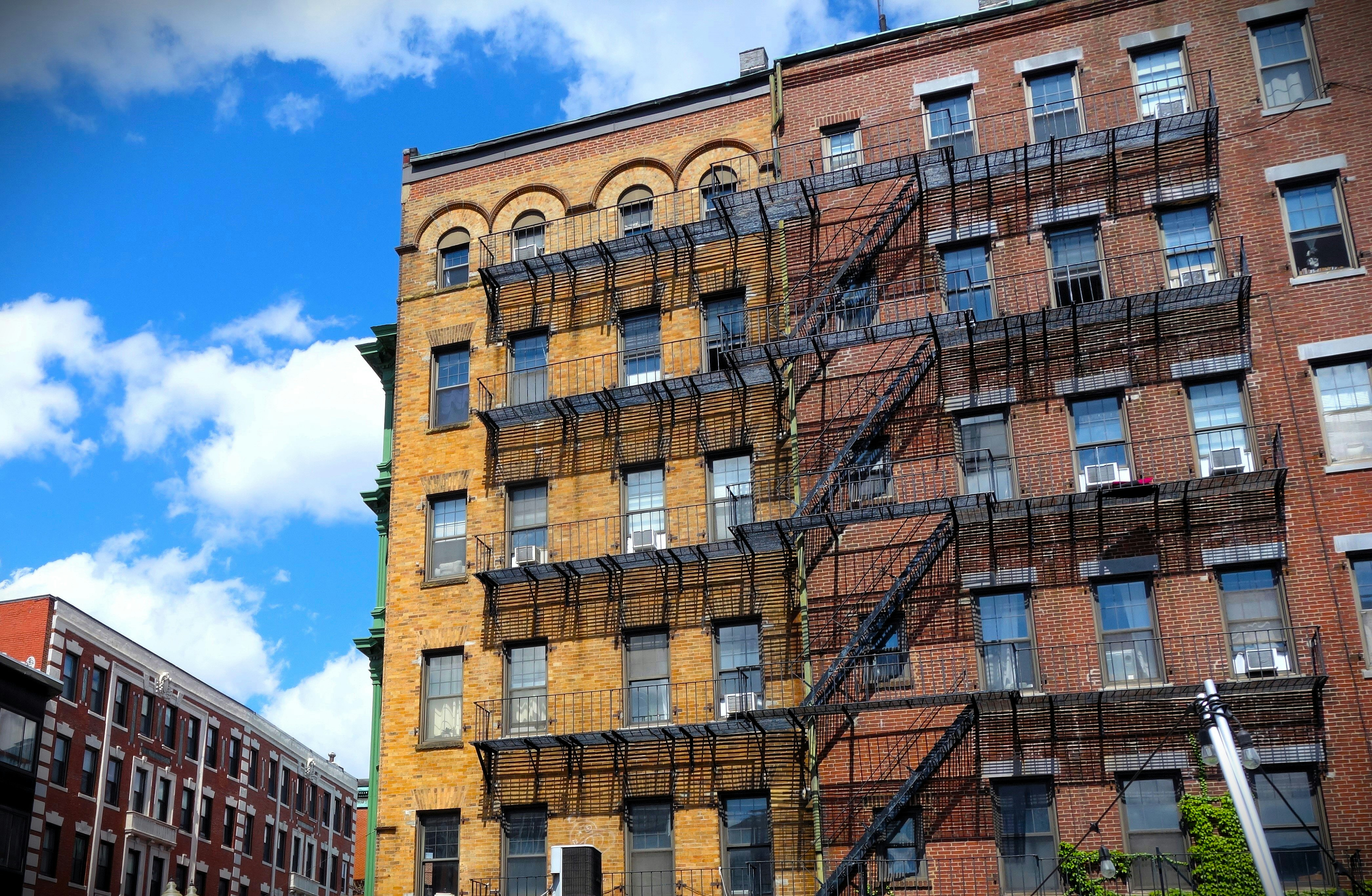 Front-facing view of a multi-story brick building with metal fire escapes and a blue sky above. A secondary building on the left frames the scene; the image captures urban architecture and textures.