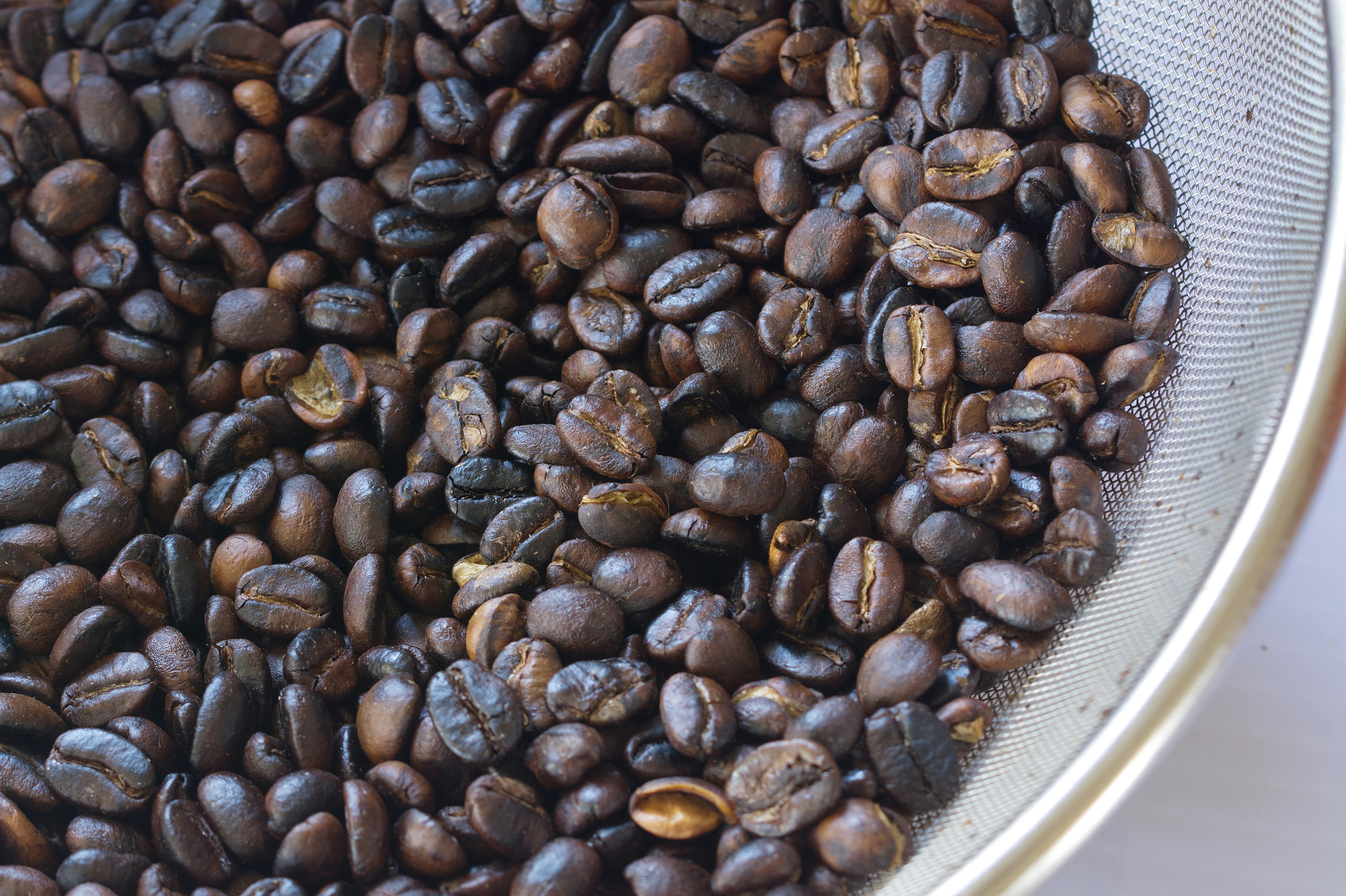 Roasted coffee beans settled in a metal sieve, revealing glossy surfaces and rich browns; the mesh and edge of the sieve hint at preparation or draining in a kitchen setting.