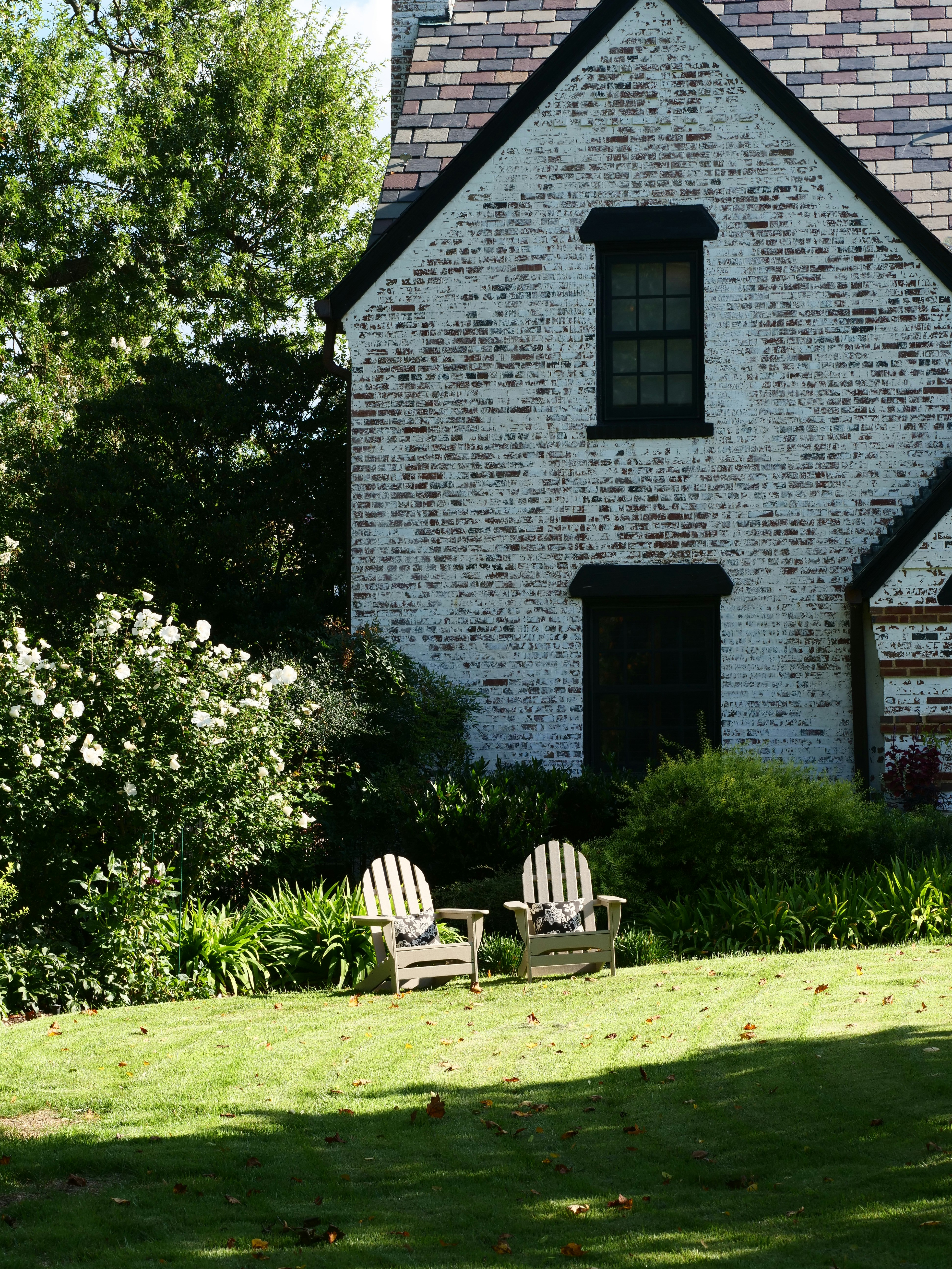 Two Adirondack Chairs in the Shade of a White Brick House Garden
