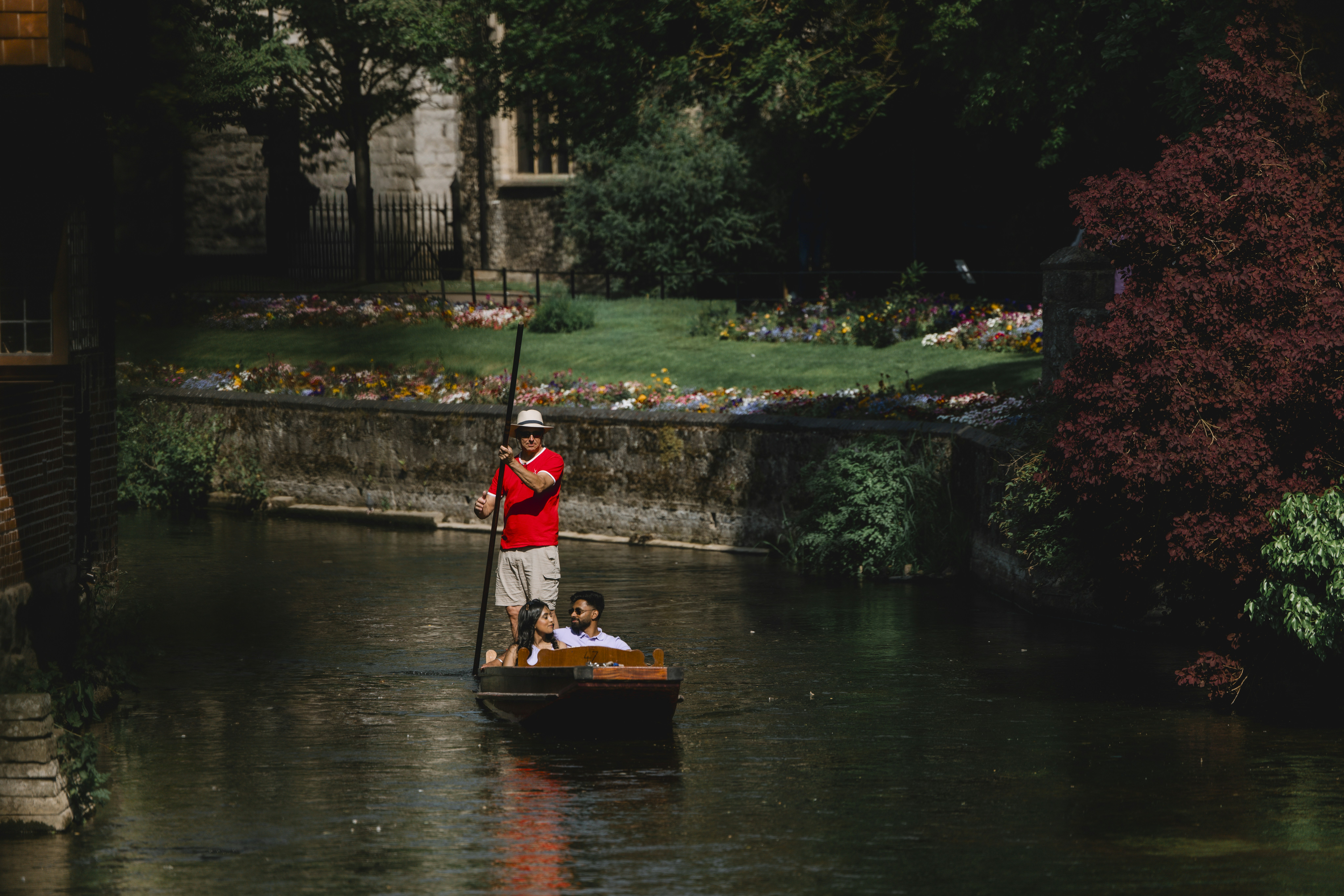 Three people ride a small boat along a shaded river beneath a stone bridge, with a red-clad spotter standing at the stern and a colorful garden lining the bank beyond the water.