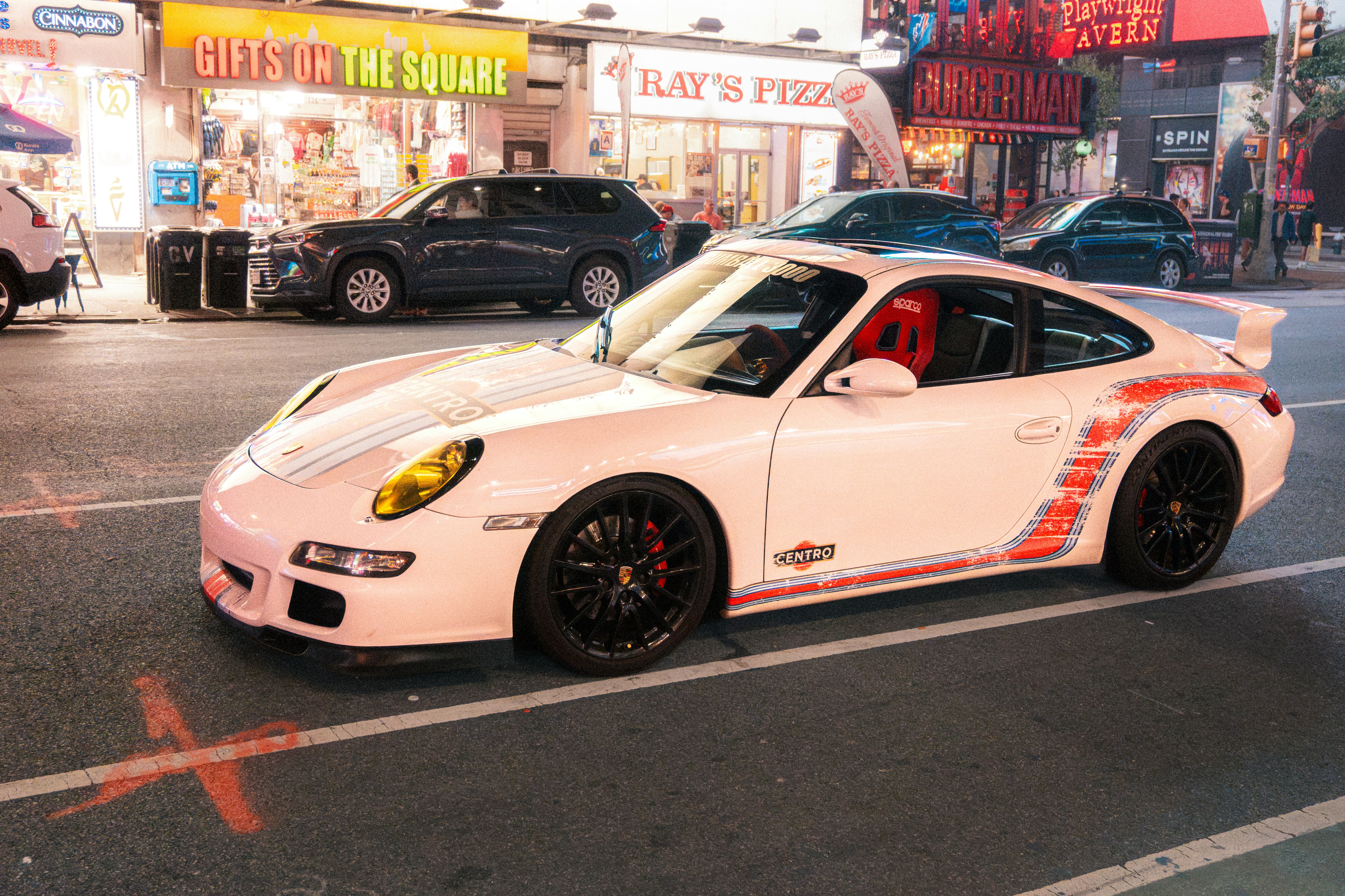 White Porsche GT3 RS with red-orange racing stripes parked on a busy city street at night; storefronts and colorful signs glow in the background.