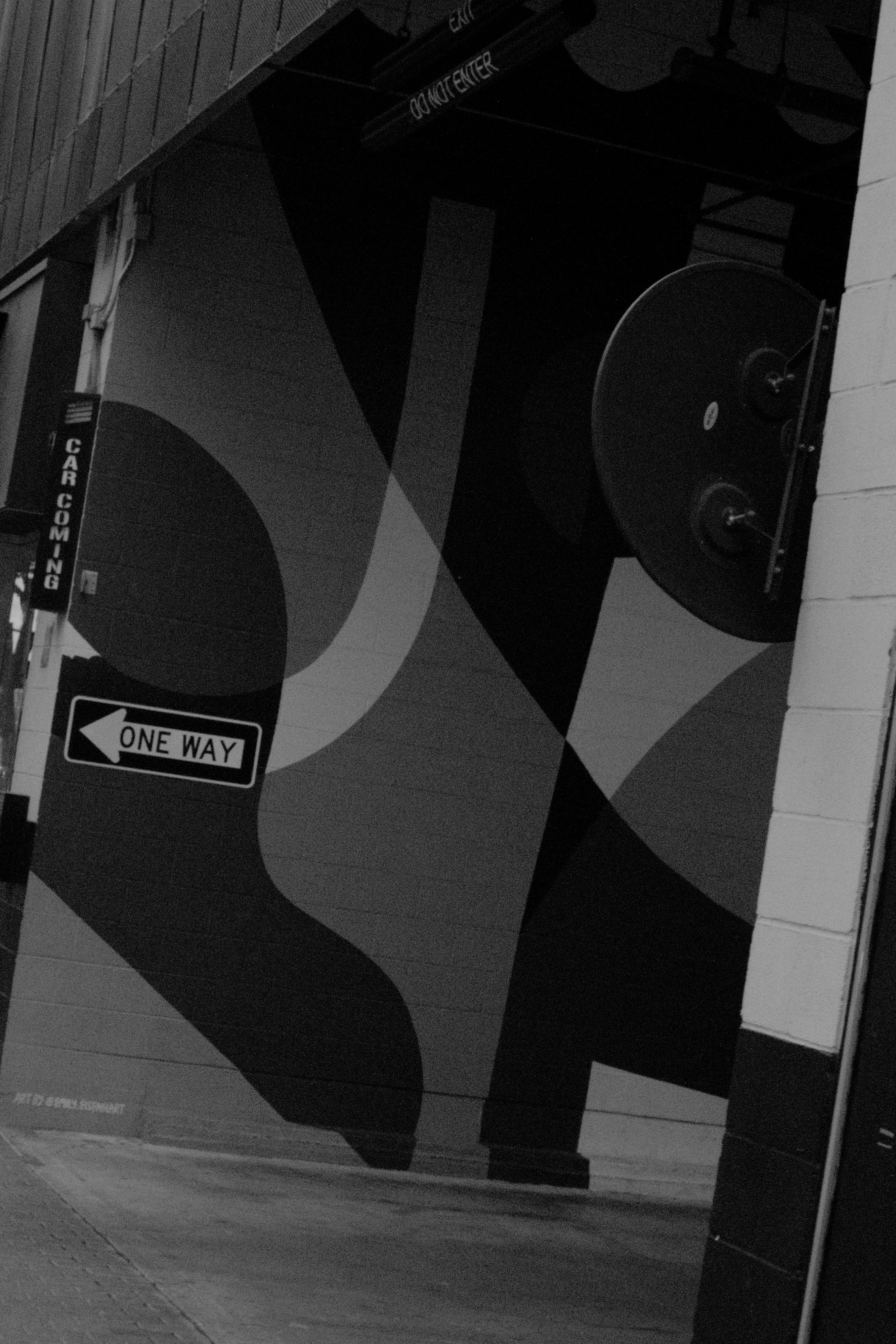 Monochrome urban mural framing a one-way sign at a corner alley; a skateboard wheel and architectural elements add depth to the composition.