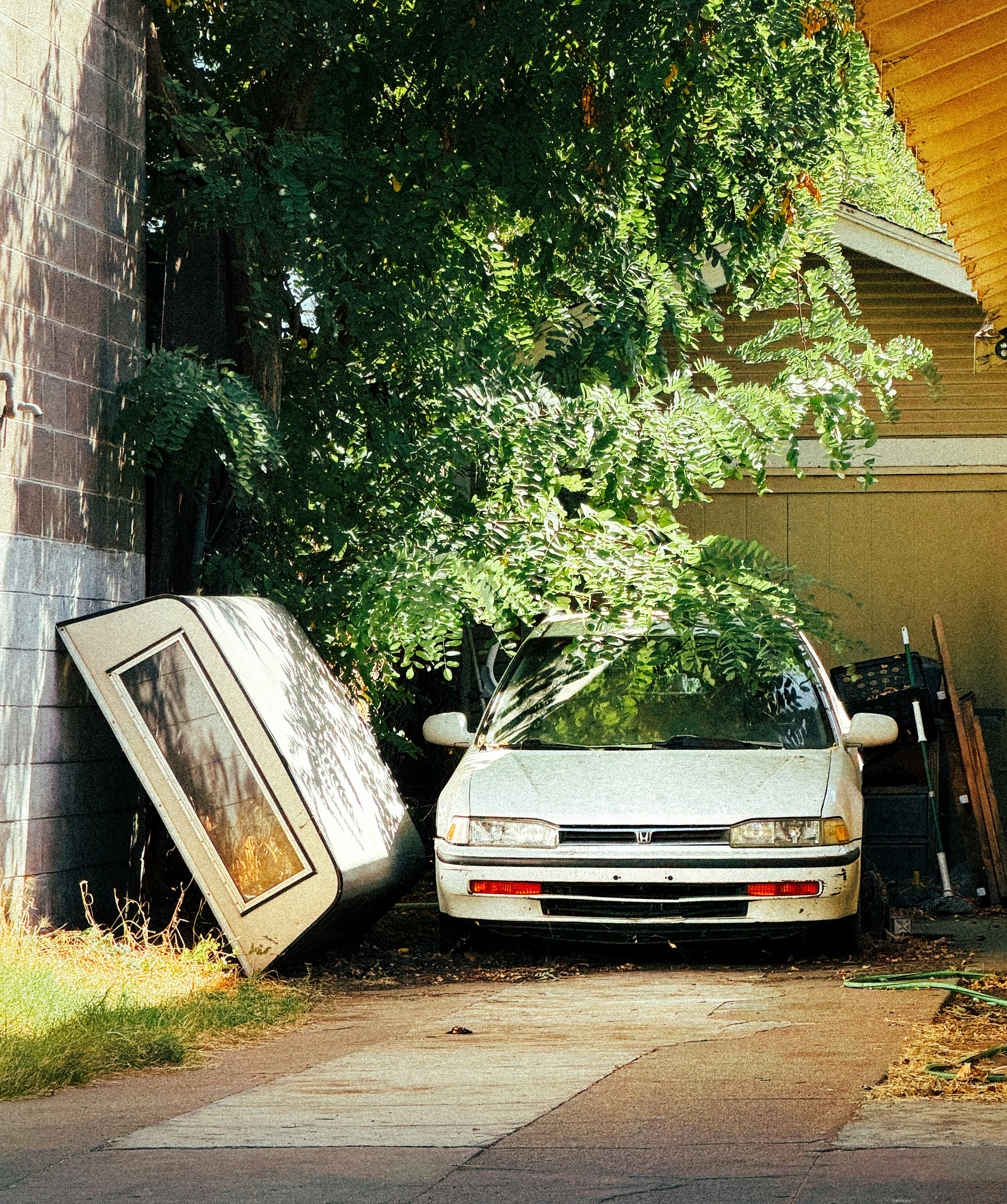 Abandoned white van under a leafy canopy, door panel and a detached cabinet leaning against a brick wall