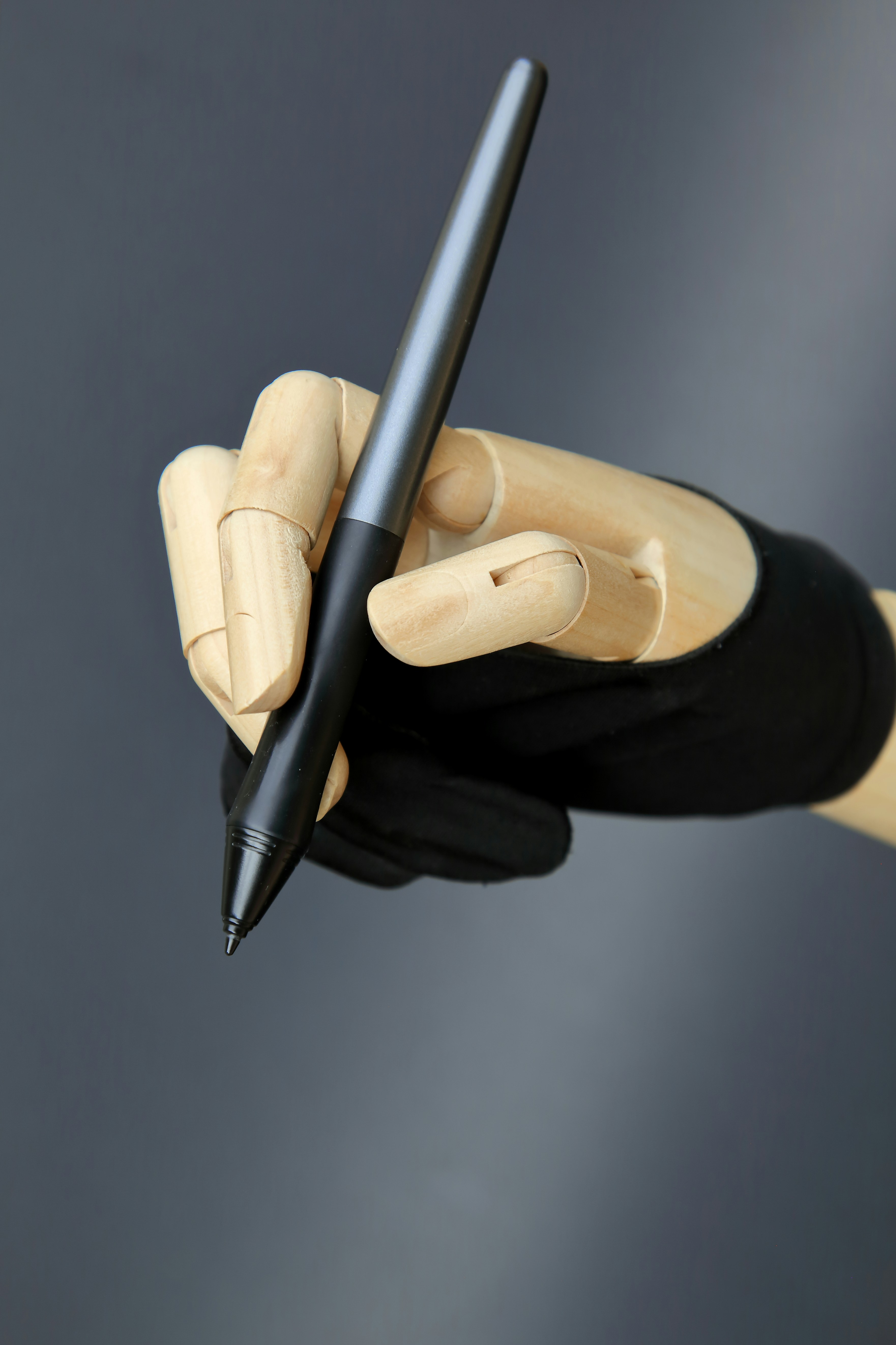 Close-up of a stylus held between wooden fingers, with a gloved hand in the background and a smooth, dark backdrop.