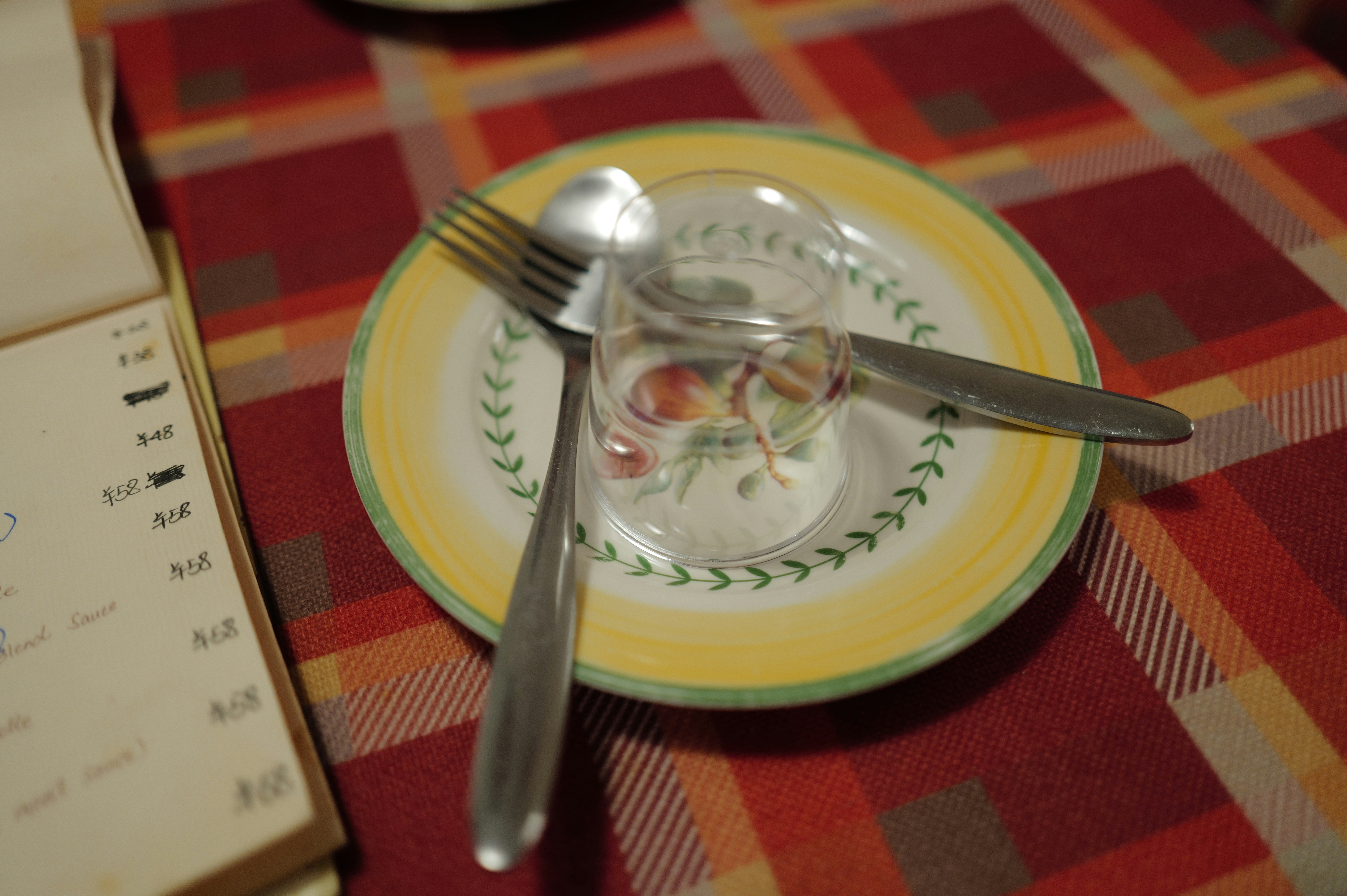 A colorful plate with a glass and cutlery rests atop a red-and-yellow checkered tablecloth, with a menu partially visible on the side.