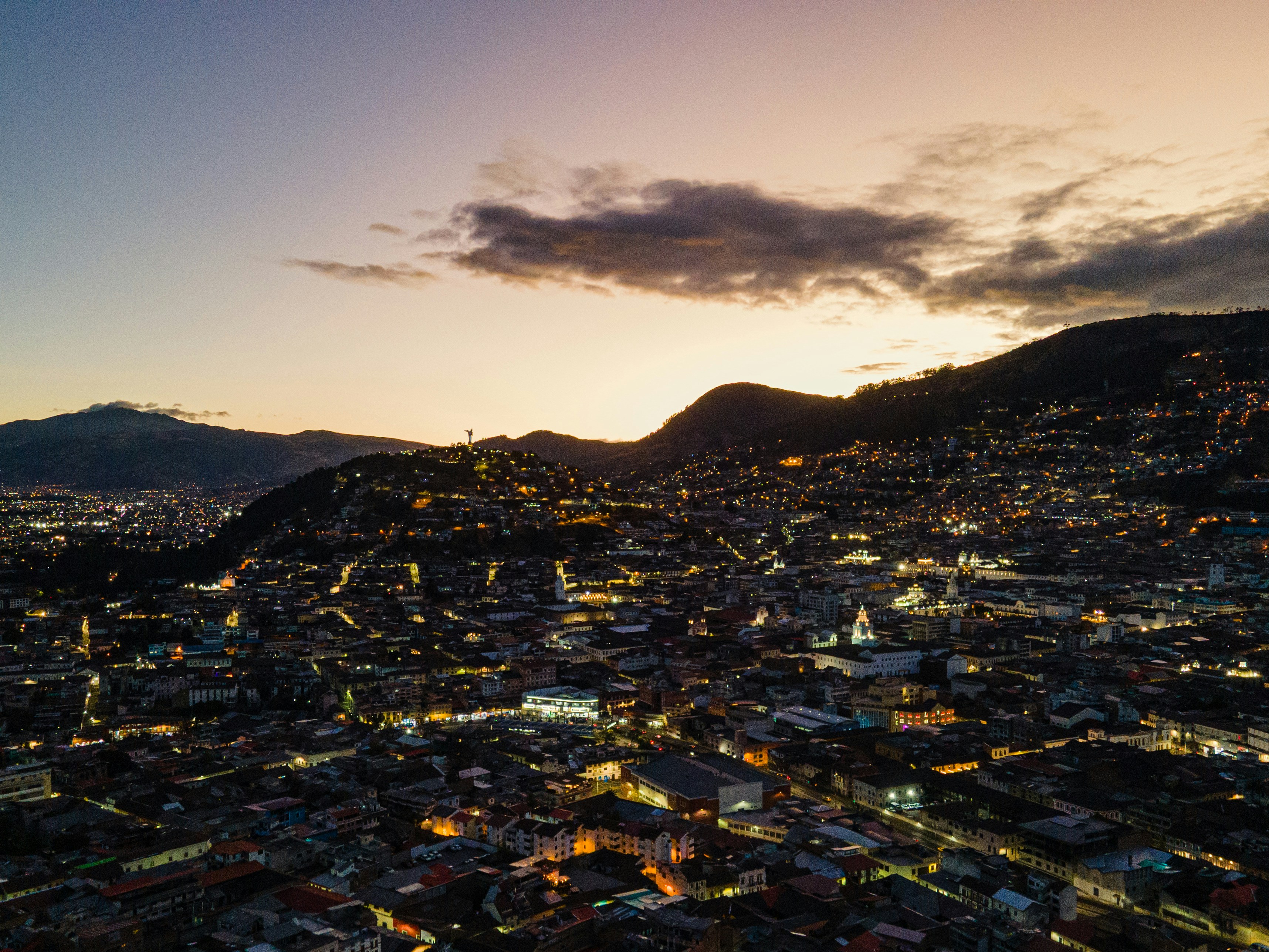 Aerial view of a densely built hillside city at dusk, lights beginning to twinkle as the sun fades; the urban grid meanders through terraced streets and low-rise rooftops. This is a nighttime cityscape capture with a drone.