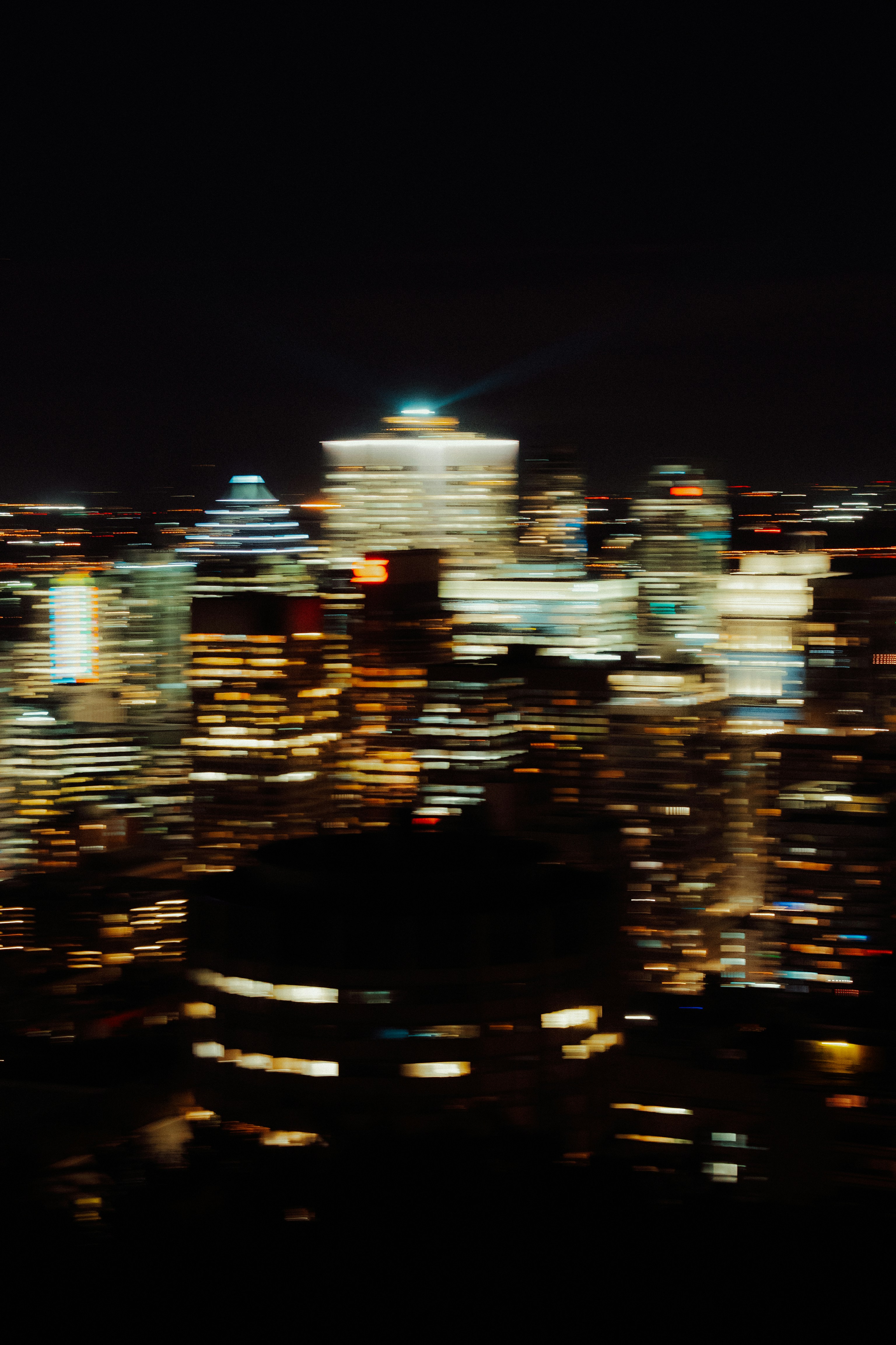 Blurred city lights at night create a painterly skyline with vertical and horizontal bokeh; the image emphasizes urban energy and motion without a central subject. It is a photographic capture of a cityscape.