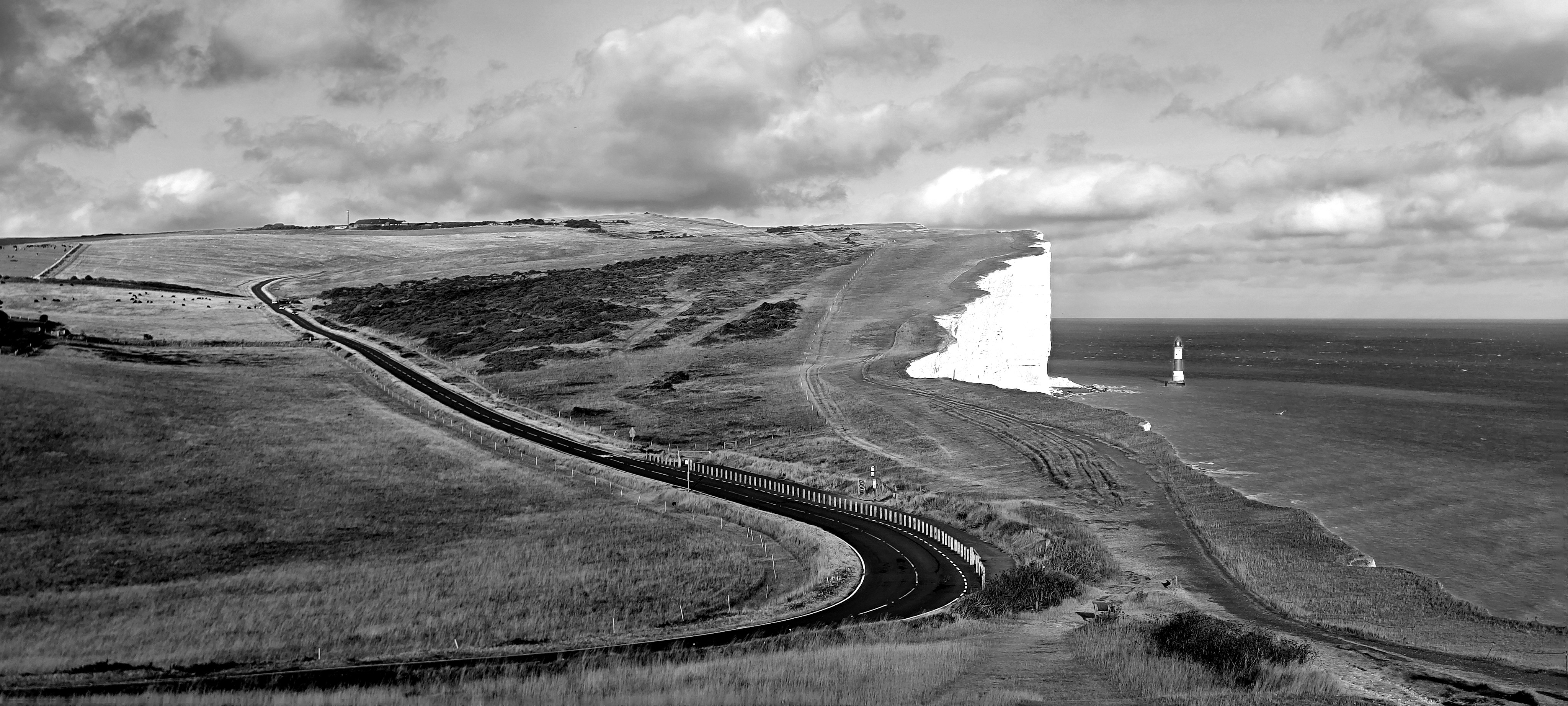 Monochrome coastal landscape featuring a winding road along grassy bluffs and towering white chalk cliffs beside the sea. The composition emphasizes vast sky, landform contours, and a distant sail under overcast light.