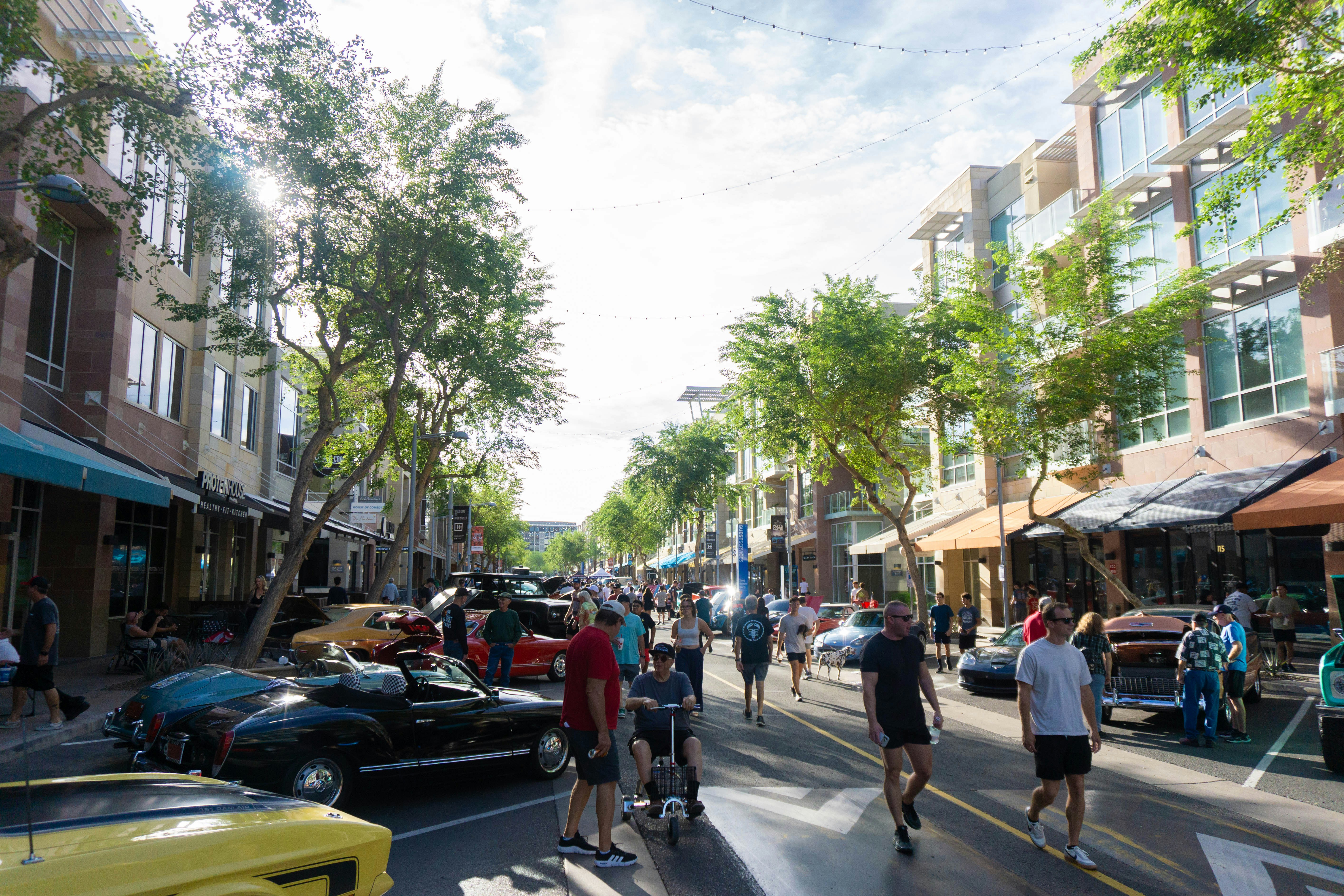 Sunlit Main Street Carnival: People, Cars, and Shops Under a Green Canopy