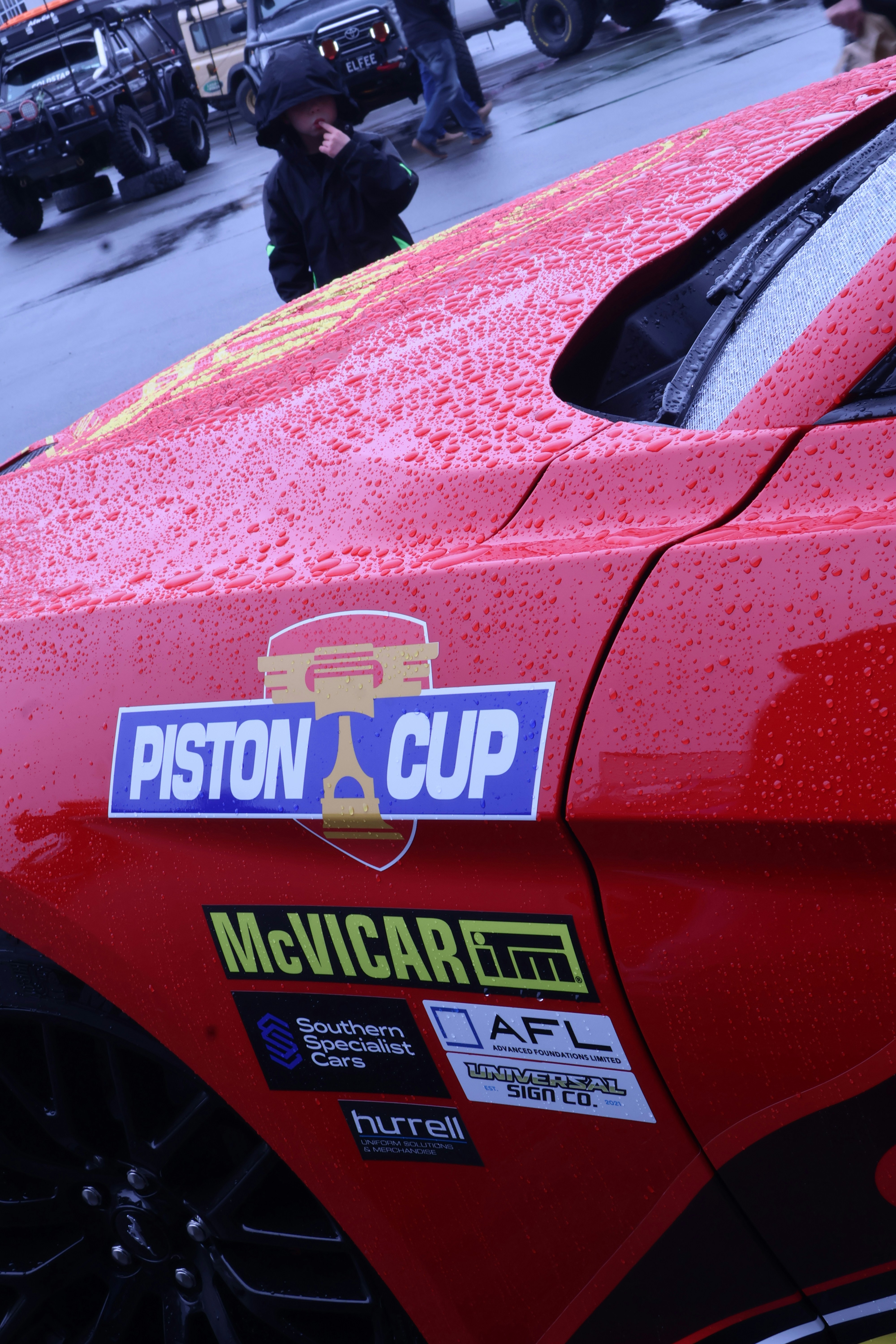 Stickered Corvette Edge: Piston Cup Racer Portrait amid Rain-Dappled Red Finish