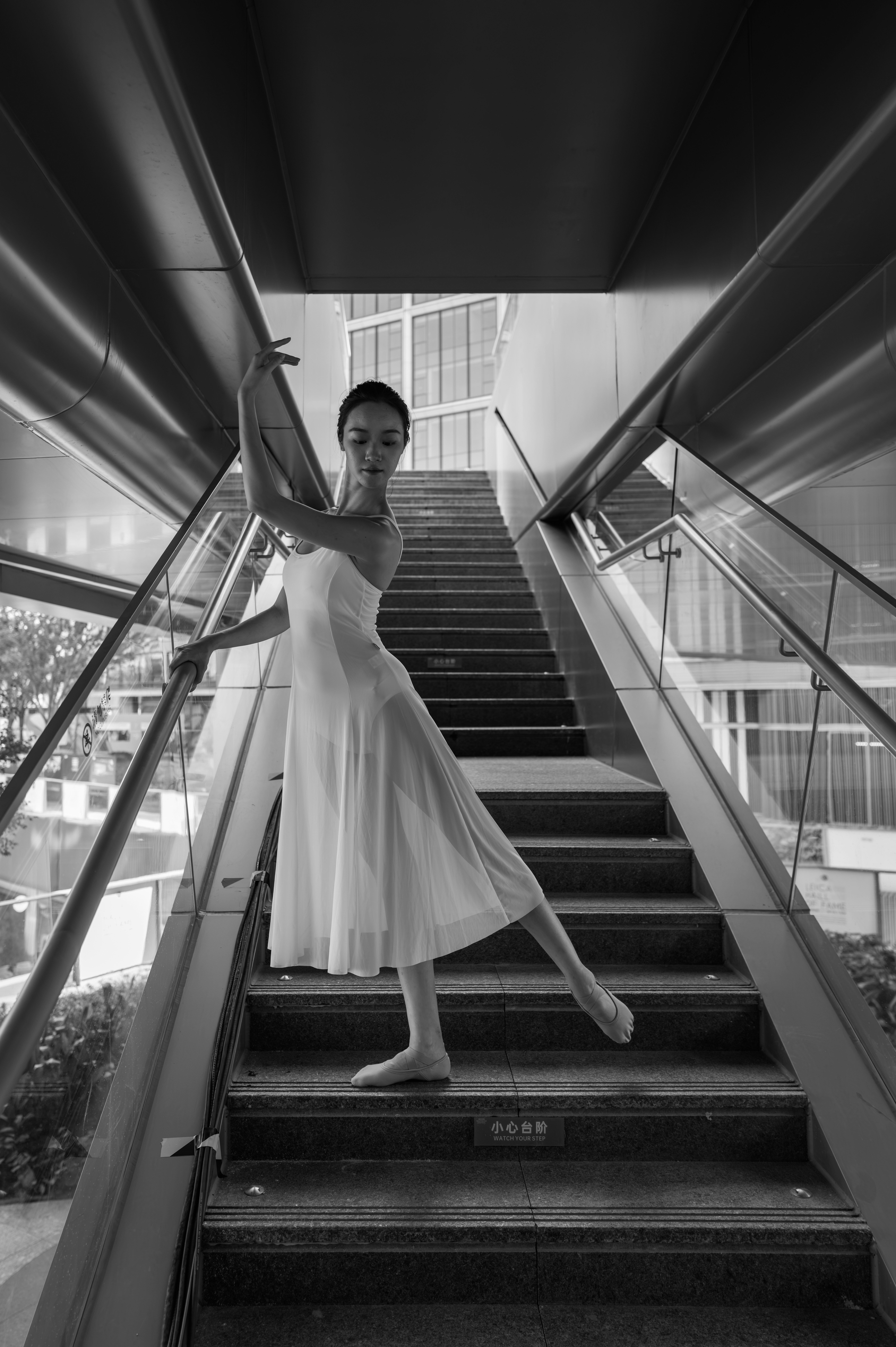 A dancer in a flowing dress poses mid-staircase in a modern, glass-walled environment; the scene emphasizes motion and form in black-and-white. This is a photographic capture, not an illustration.