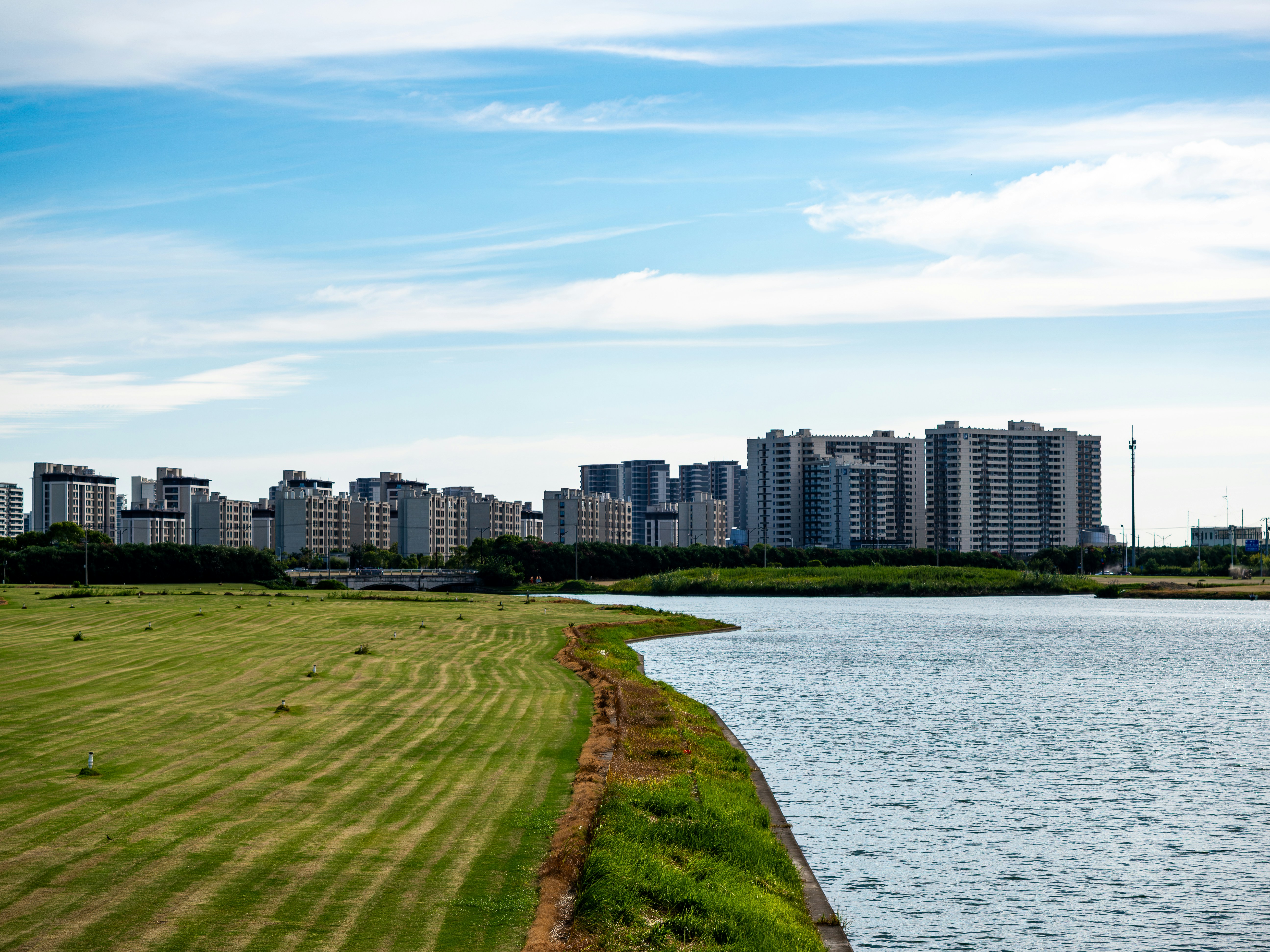 A grassy riverside path curves toward a distant cityscape along the water's edge. The scene juxtaposes wide, open foreground with a dense row of modern buildings across the river.