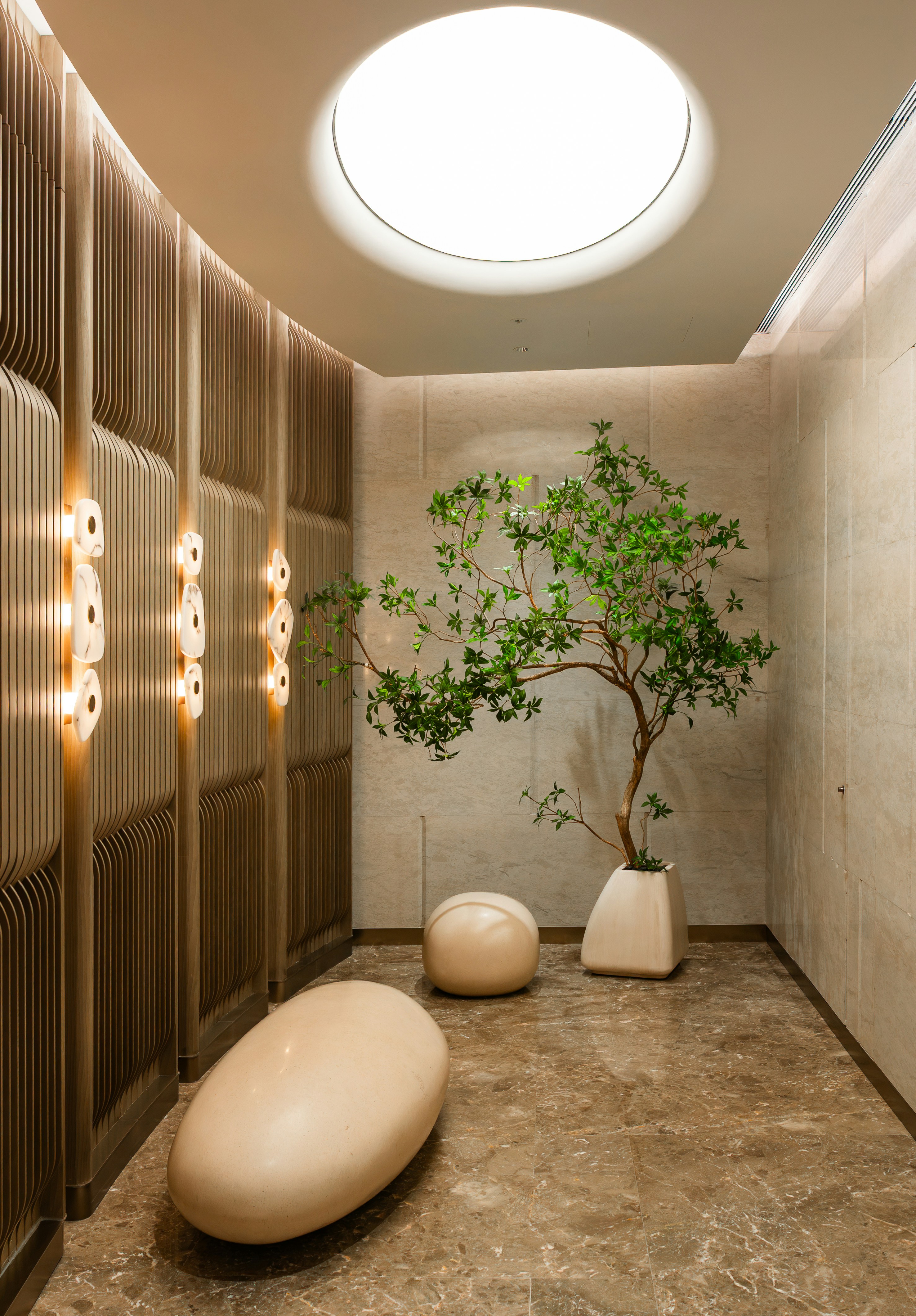 A modern interior scene features a small tree in a white planter and soft sculptural seating along a marble floor, with warm wall lighting and architectural panels in the background.