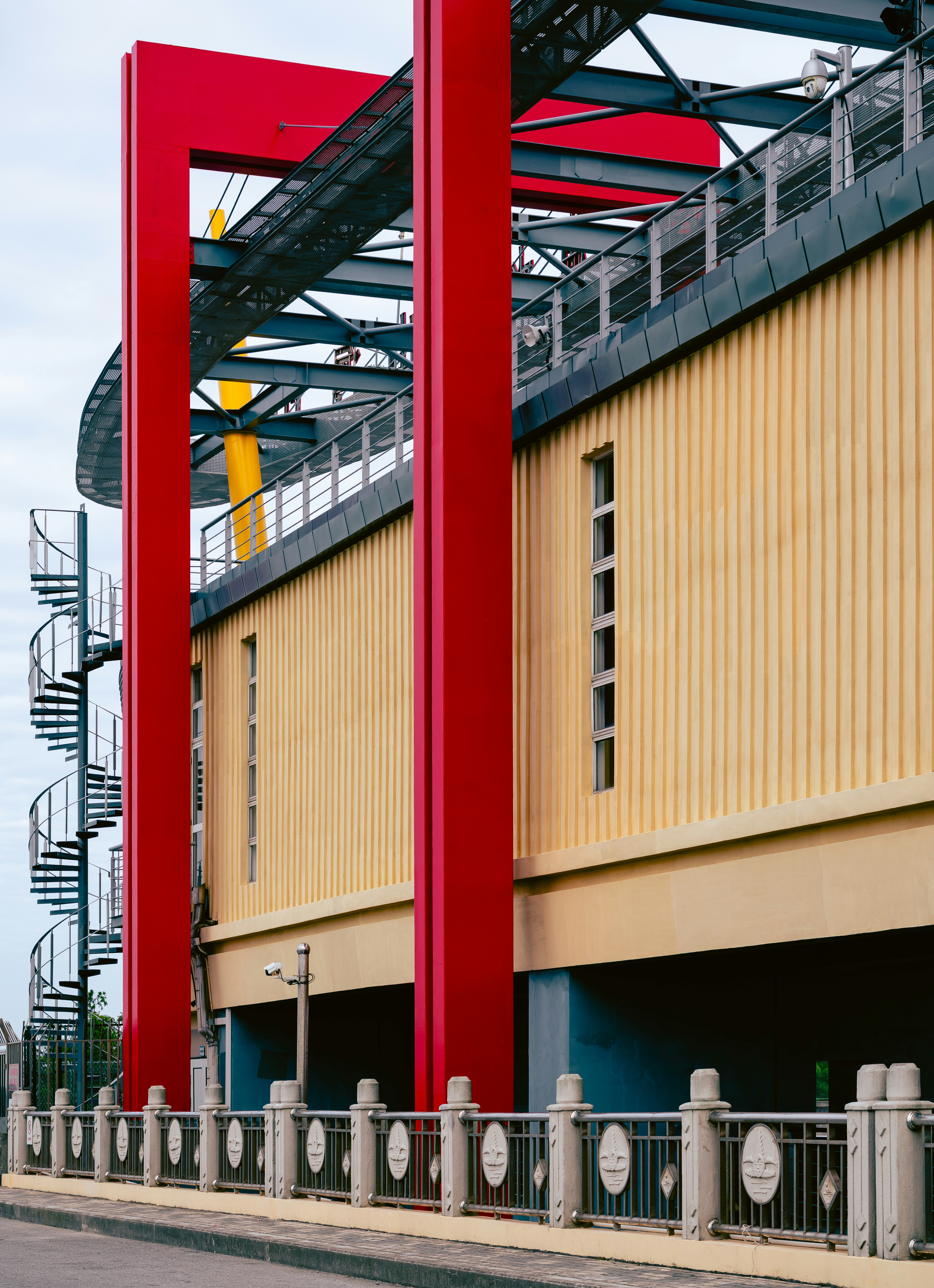 Bold red steel frames cut through a sunlit industrial building with corrugated yellow walls and a pedestrian railing; architectural lines lead the eye along the structure.