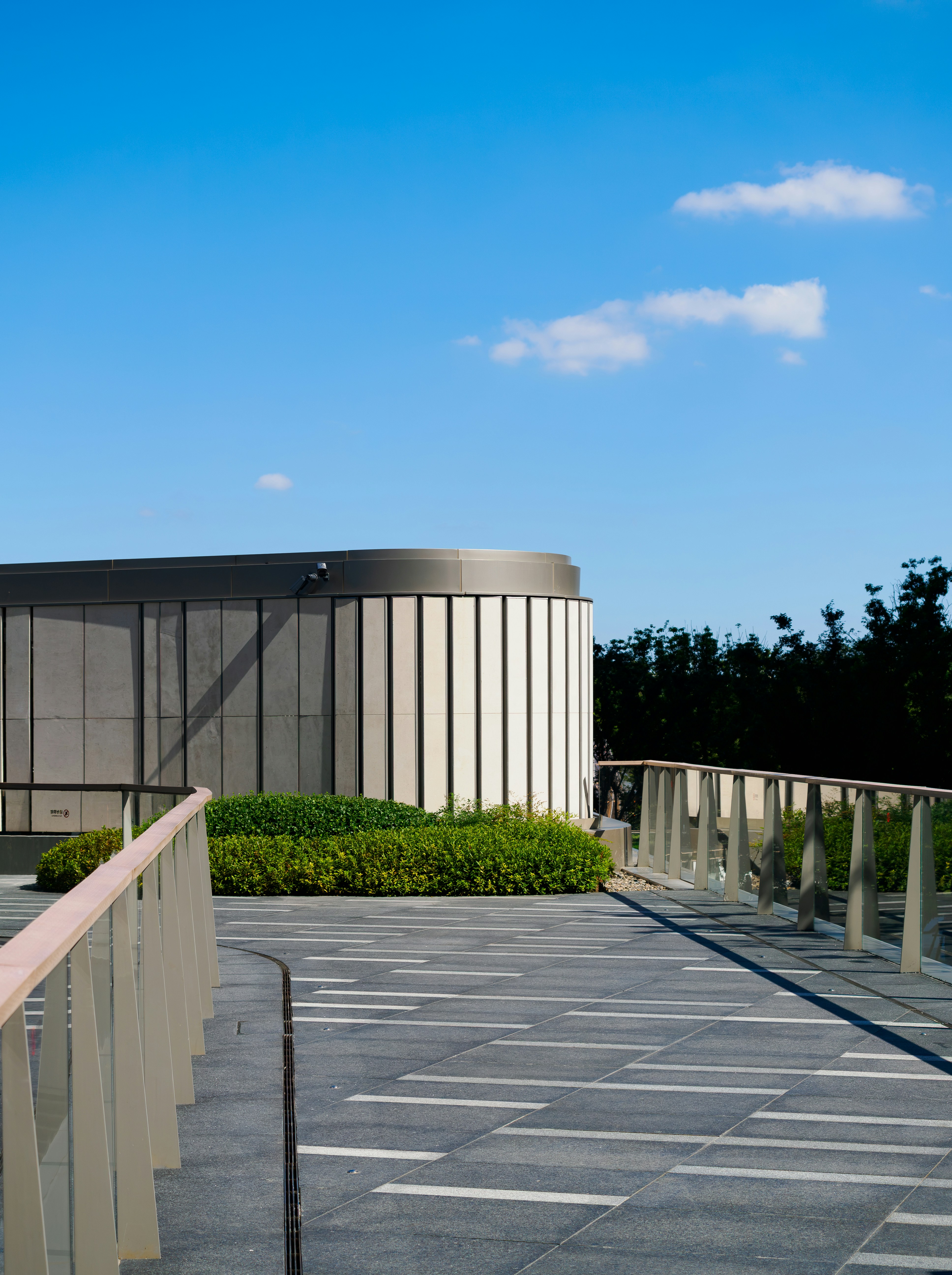 Sculpted modern rooftop walkway leads to a rounded architectural annex with a bright blue sky above; the scene emphasizes geometric lines and trimmed greenery.