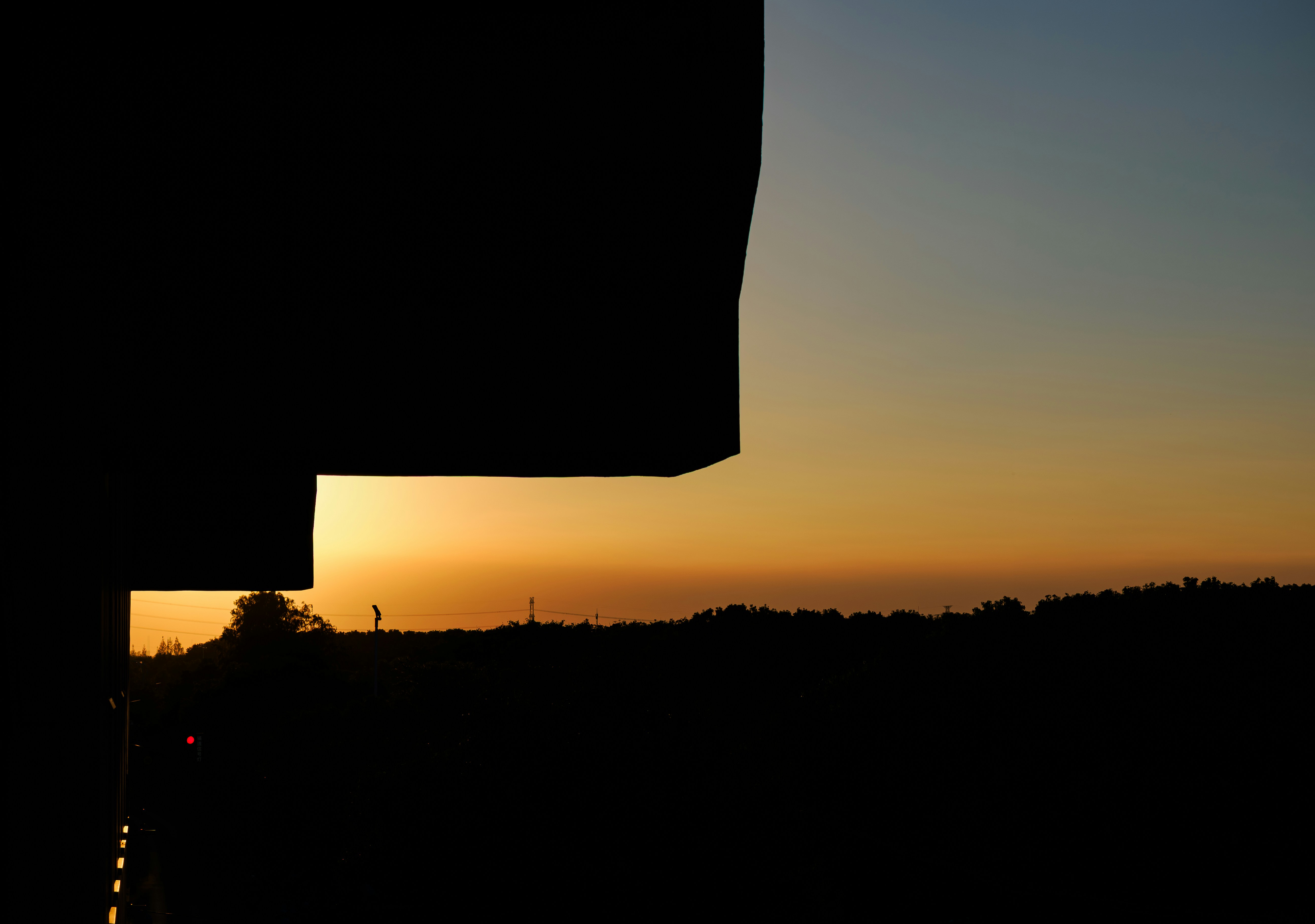 Silhouetted overhang against a warm sunset horizon, with distant treeline and subtle city lights; a minimal architectural shape dominates the left, emphasizing color gradient in the sky. (DSLR-like silhouette scene)