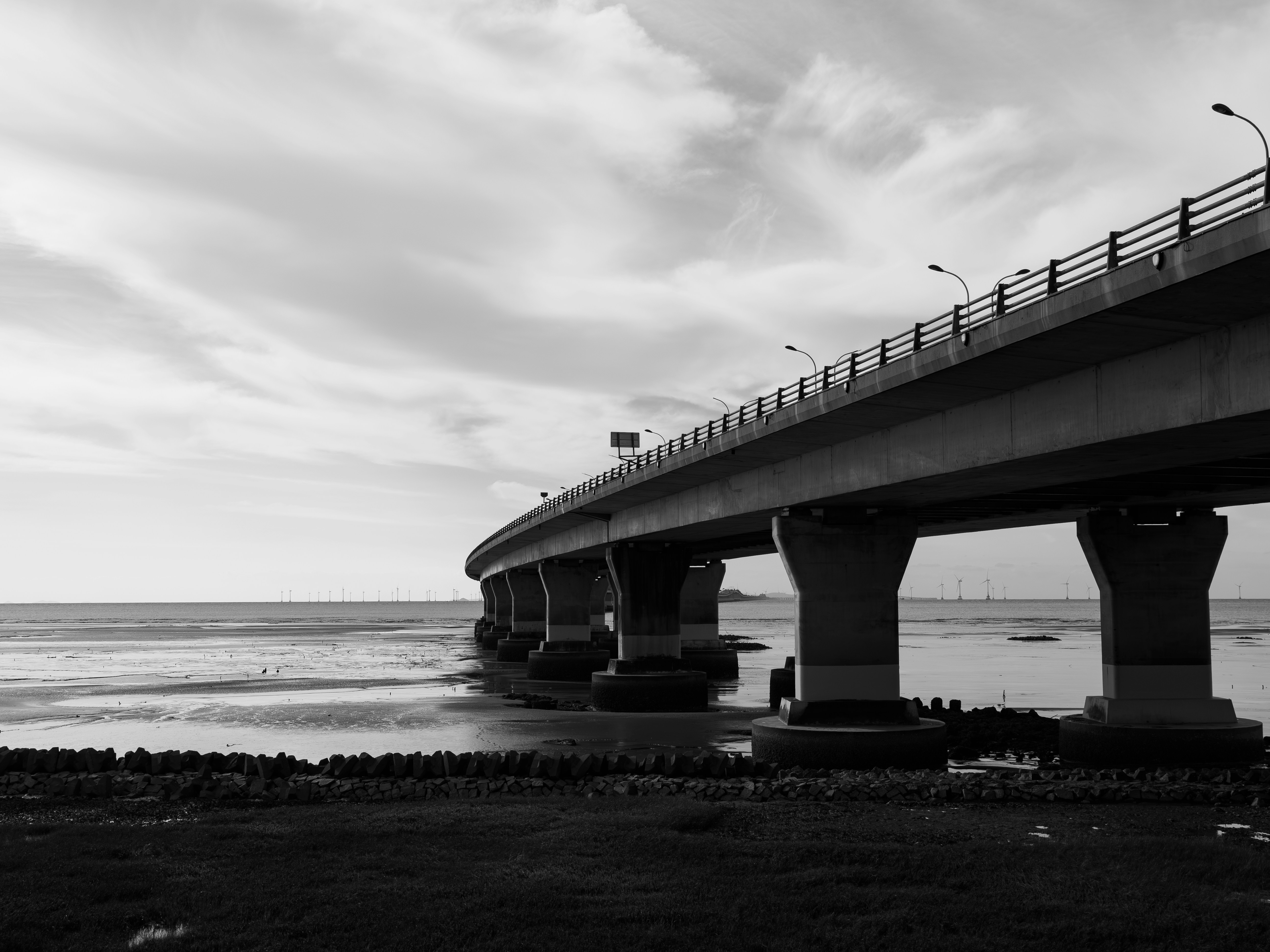 Monochrome view of a seaside bridge extending from the right, its supporting piers marching toward the horizon where the calm water meets the sky.