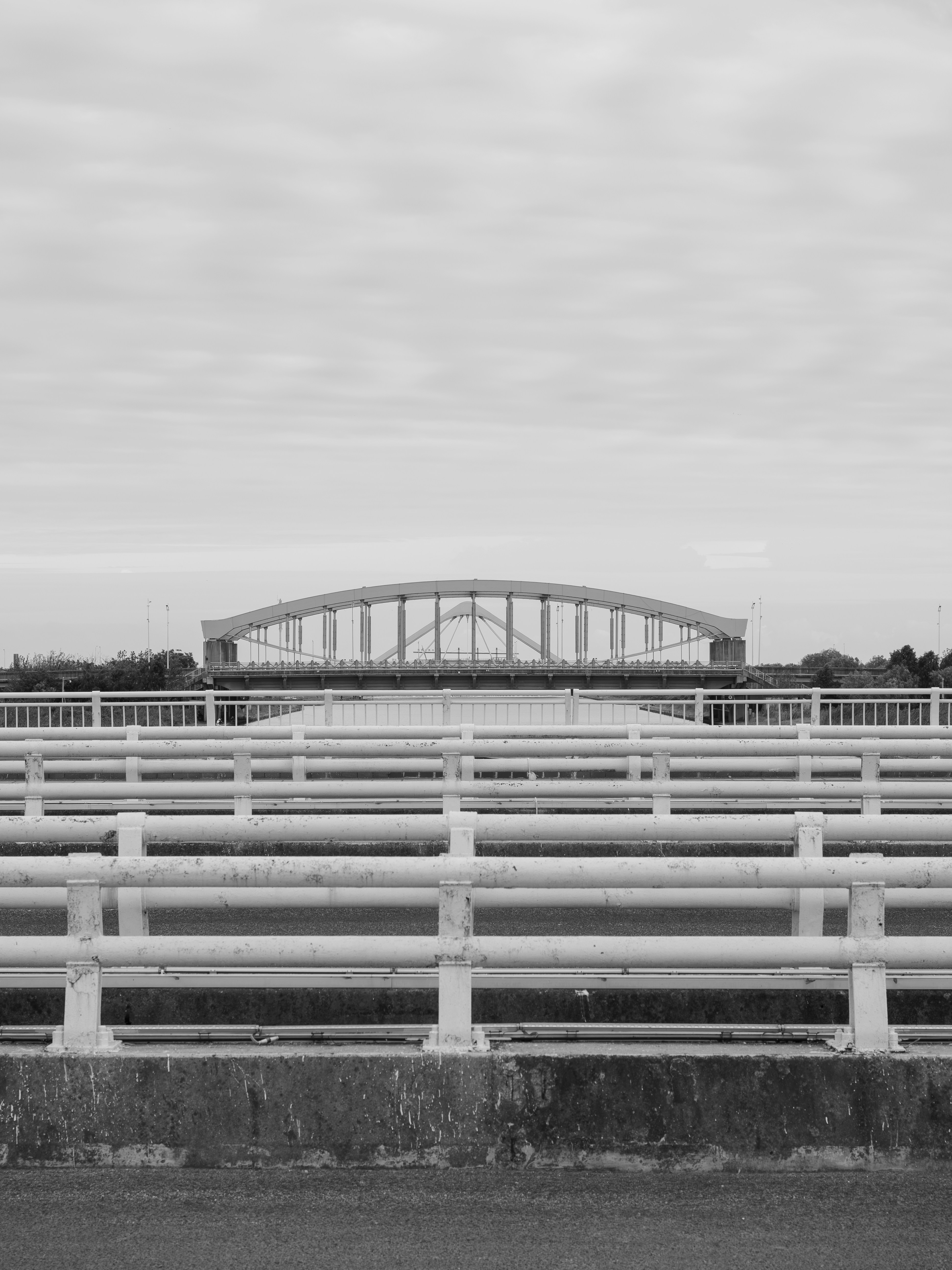 Monochrome scene of a distant arched bridge seen over a foreground of stacked white guard rails, conveying depth and symmetry.