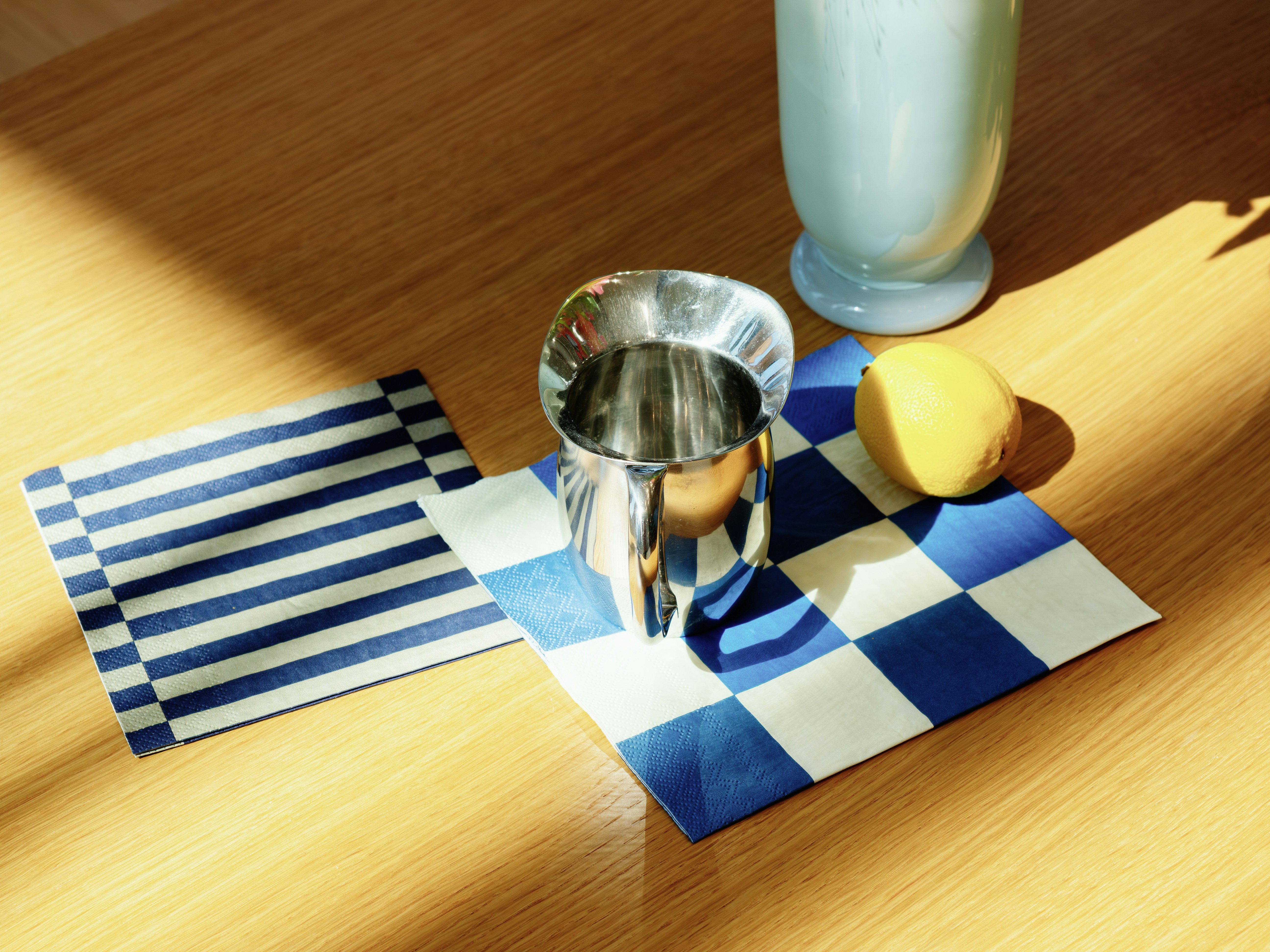 A sunlit tabletop still life featuring a striped fabric placemat, a folded checkered cloth, a shiny metal pitcher, a pale-blue drinkable vessel, and a bright lemon casting warm reflections on the wooden surface.
