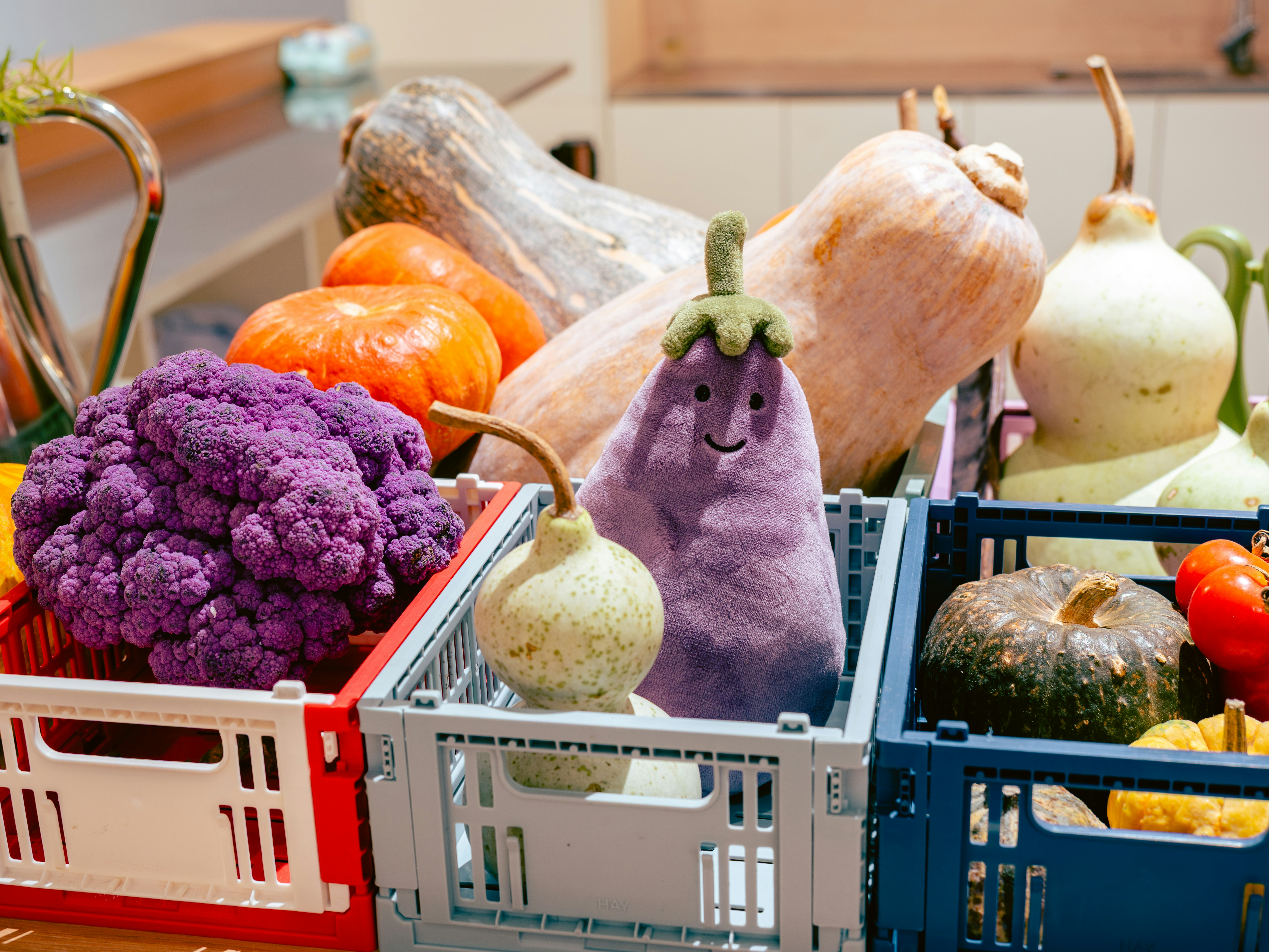 Colorful vegetables and a plush eggplant toy sit among crates of produce, creating a playful still-life in a market or kitchen setting.