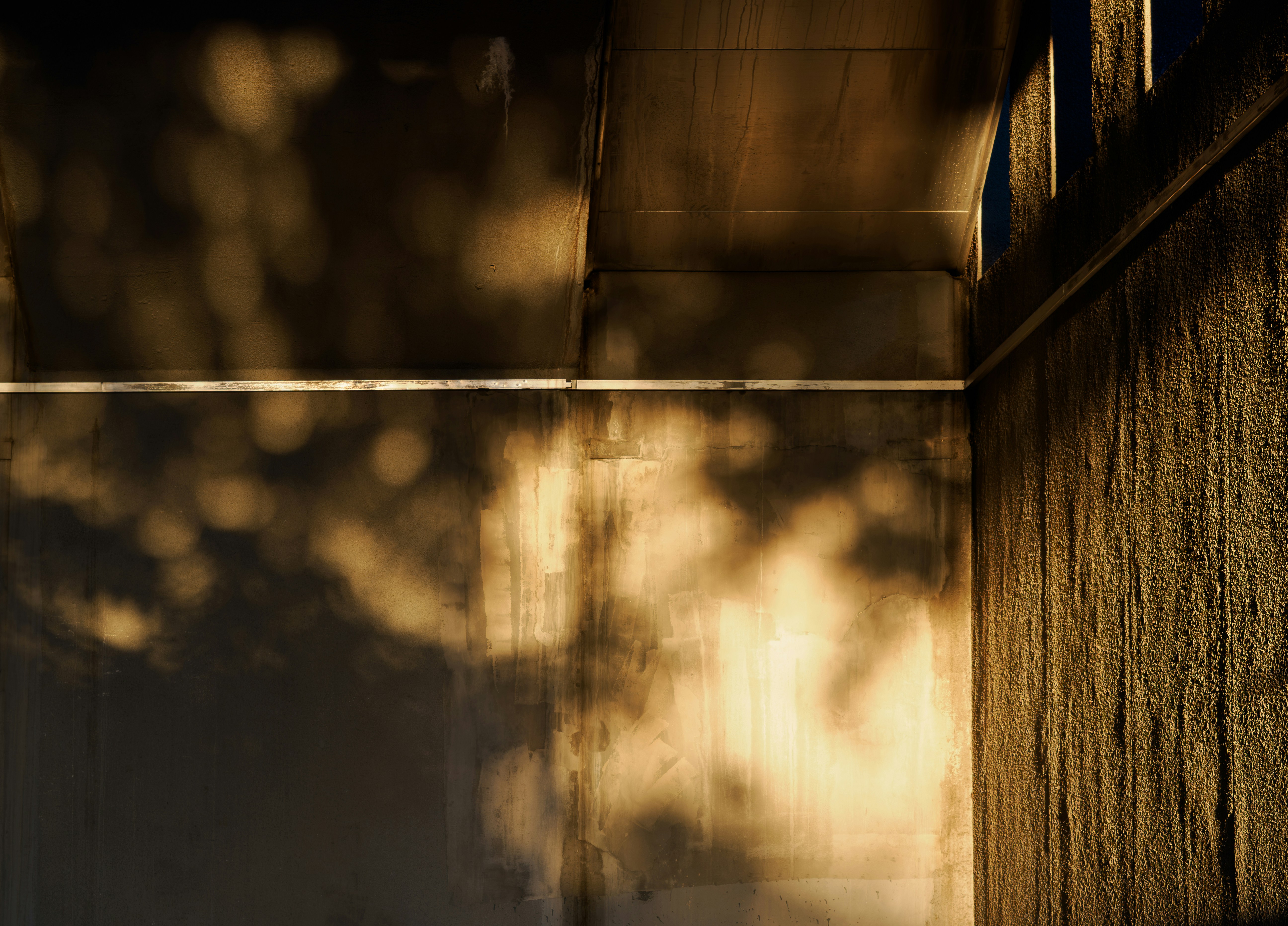 Warm, moody corner of an urban structure bathed in soft golden light, with textured concrete walls and a beam of sun-dappled highlight creating abstract patterns.
