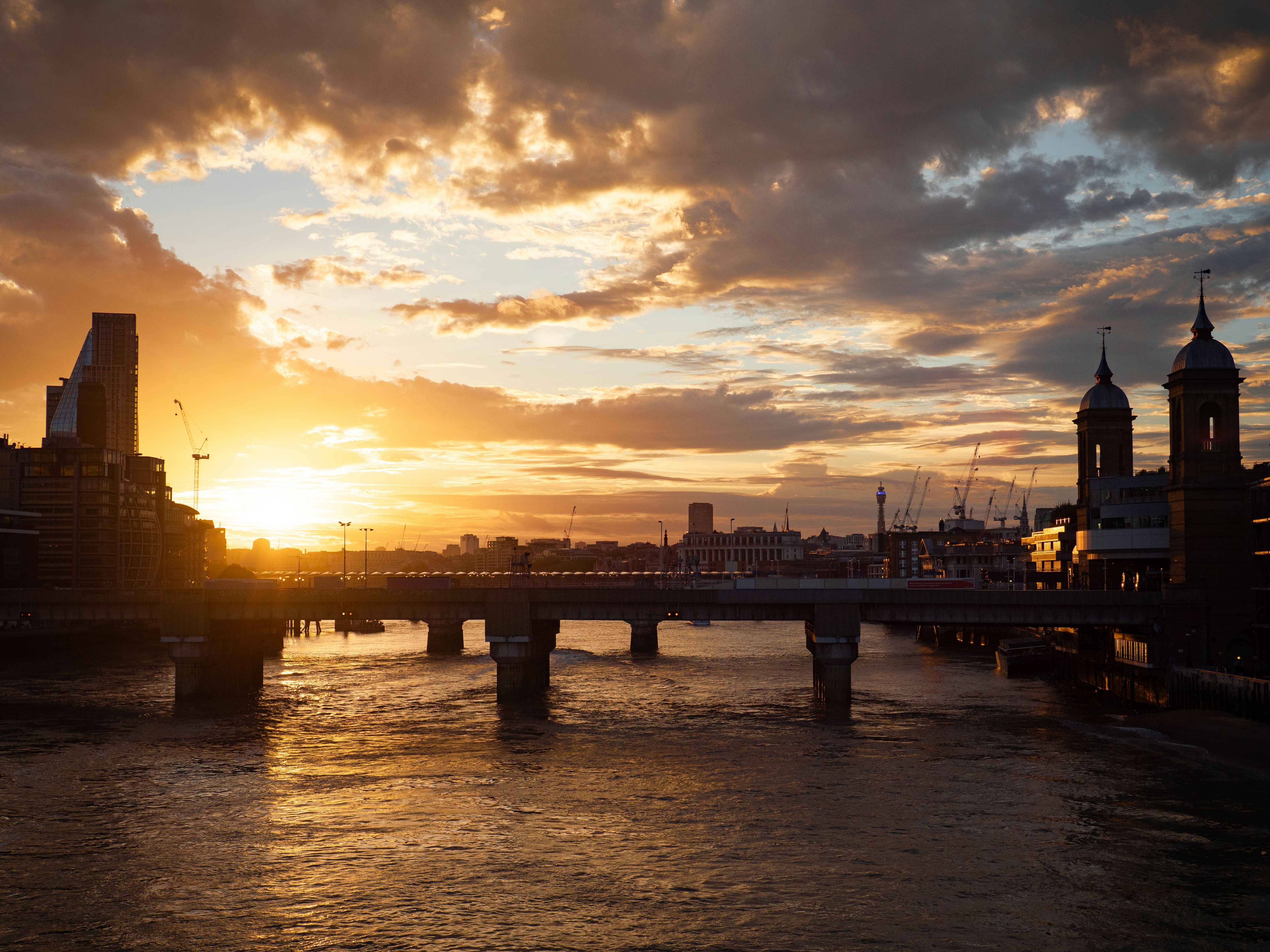 Rising sun over a city river with a bridge silhouette and warm clouds; architectural towers frame the glow on the right, water reflecting the sky, suggesting a calm morning commute.