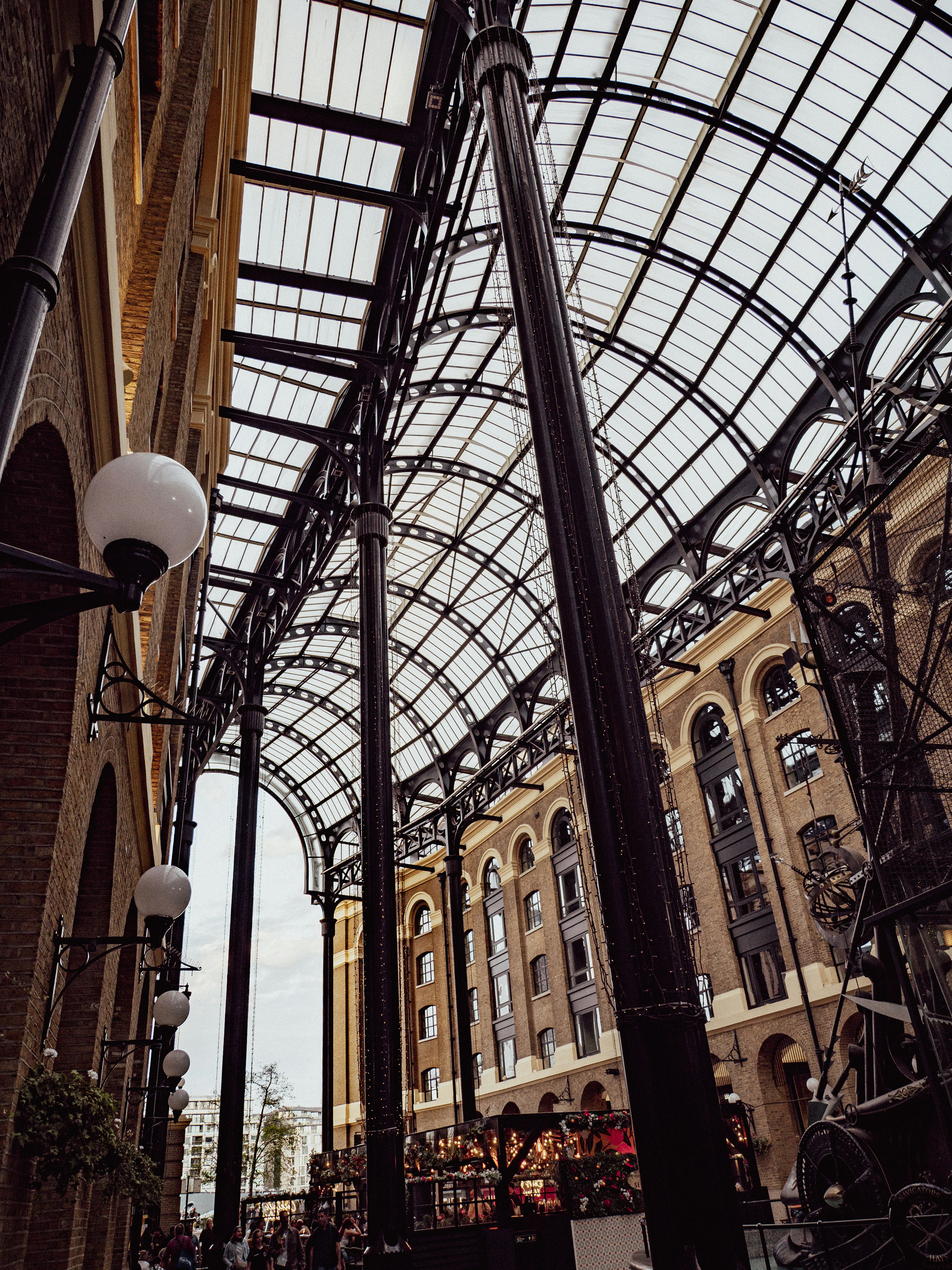 Indoor urban atrium with tall black iron columns and a curved glass roof, pedestrians at the lower level, warm brick walls and decorative lamps.