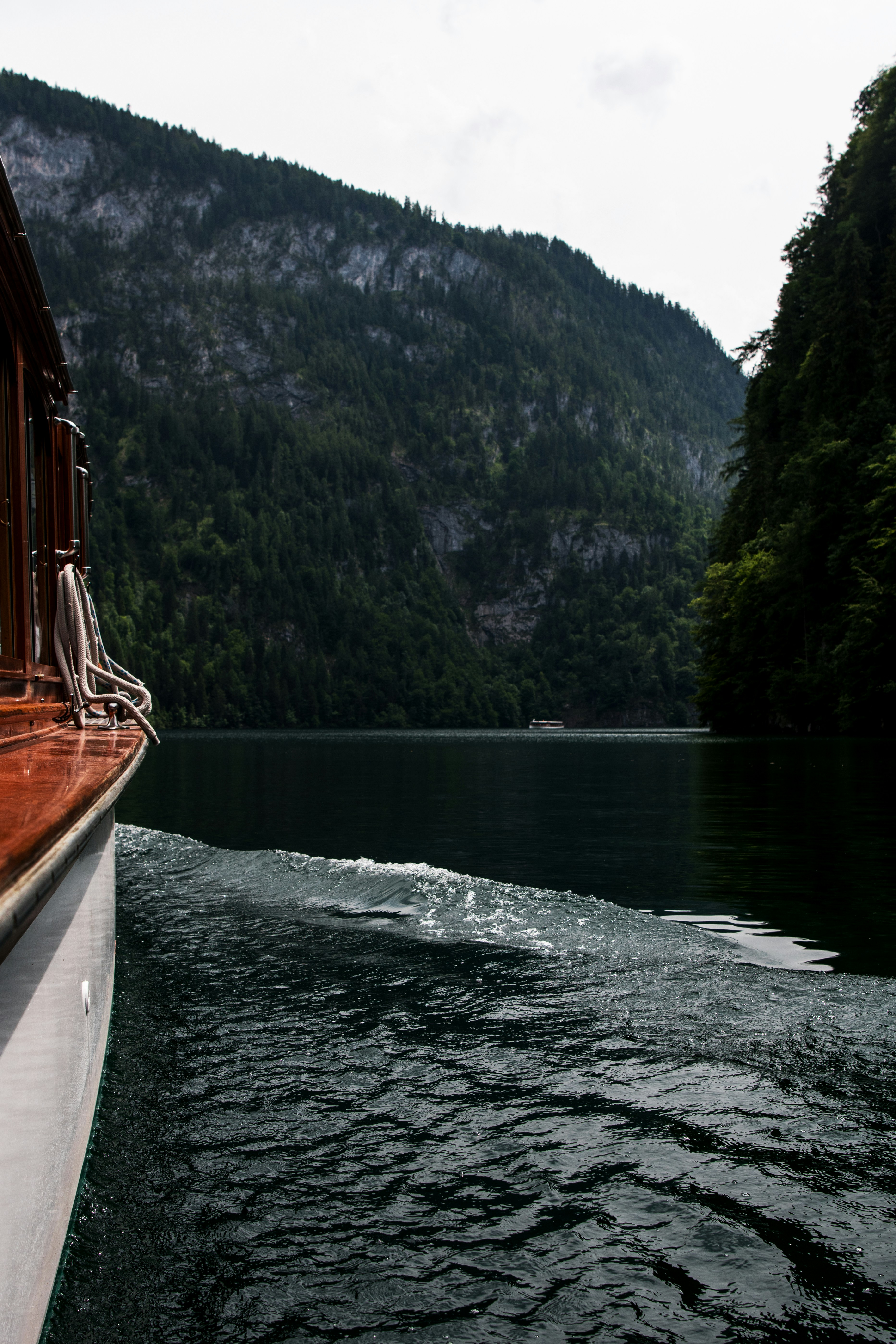 Wooden boat glides along a calm alpine lake with dark forested cliffs; distant wake hints at motion and a tiny boat on the far shore adds scale.