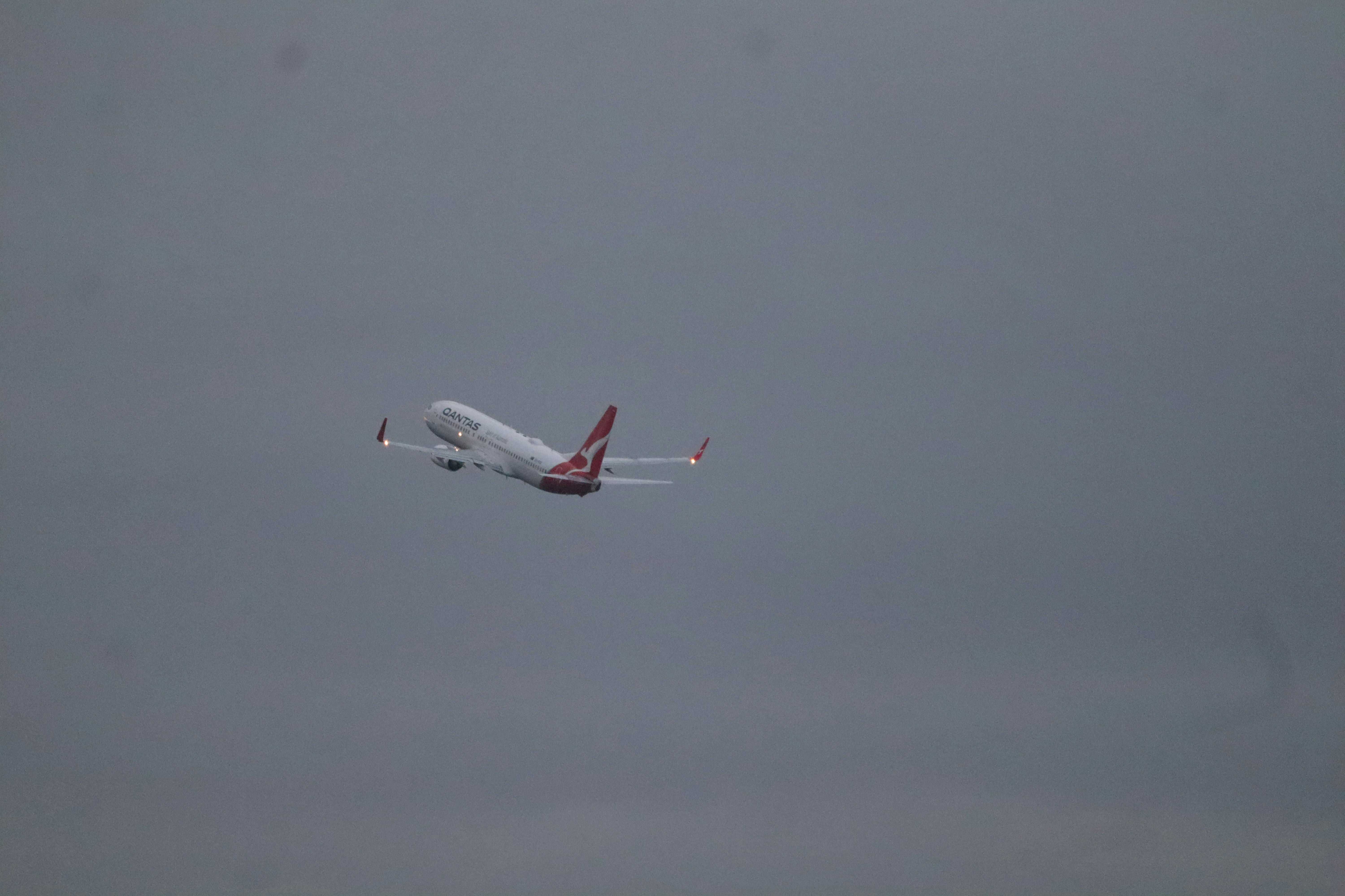 A distant passenger jet climbs against a gray, overcast sky, its red tail visible as the main focal element.