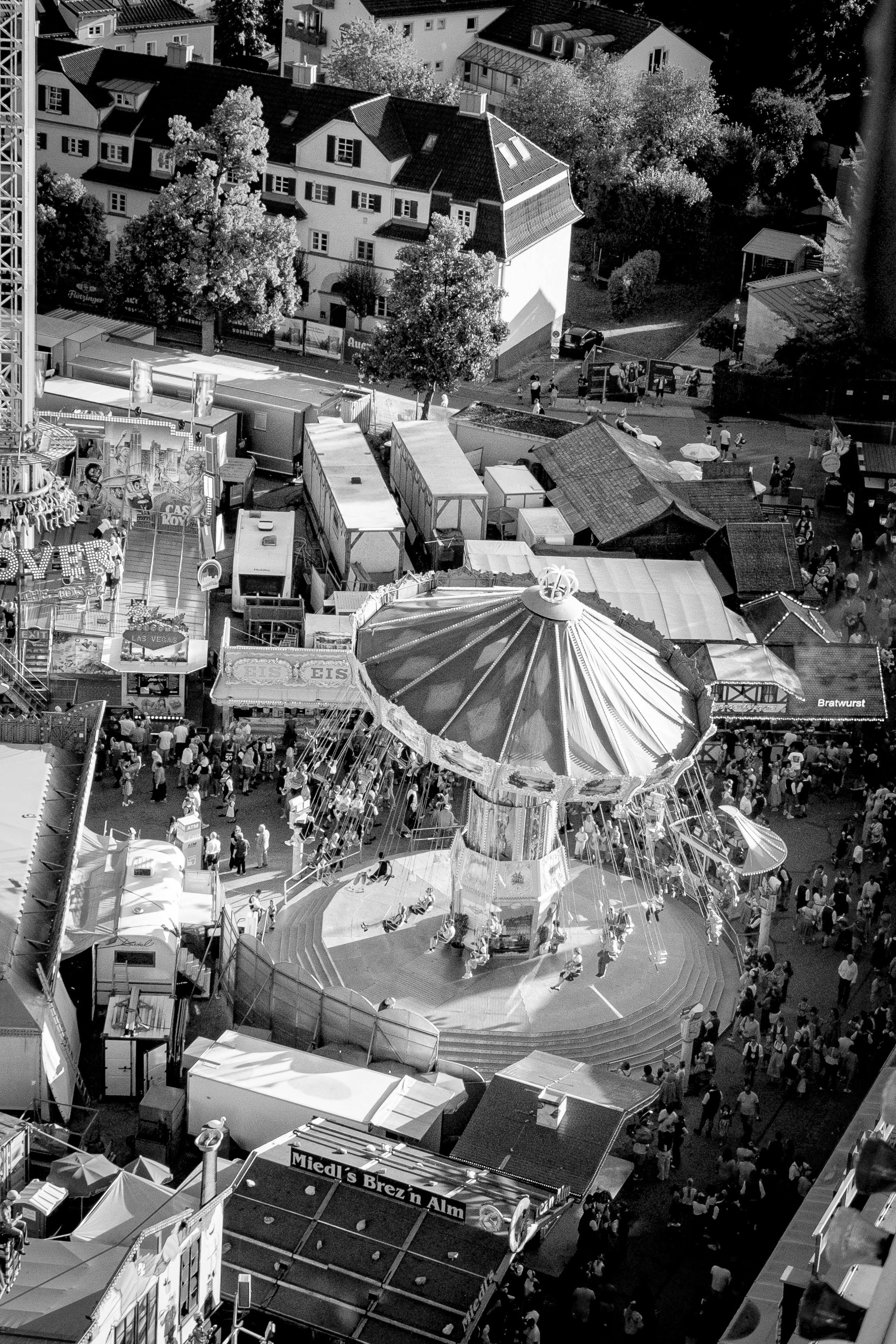 A bird’s-eye view of a bustling carnival with a grand carousel at the center, surrounded by tents, stalls, and a dense crowd.