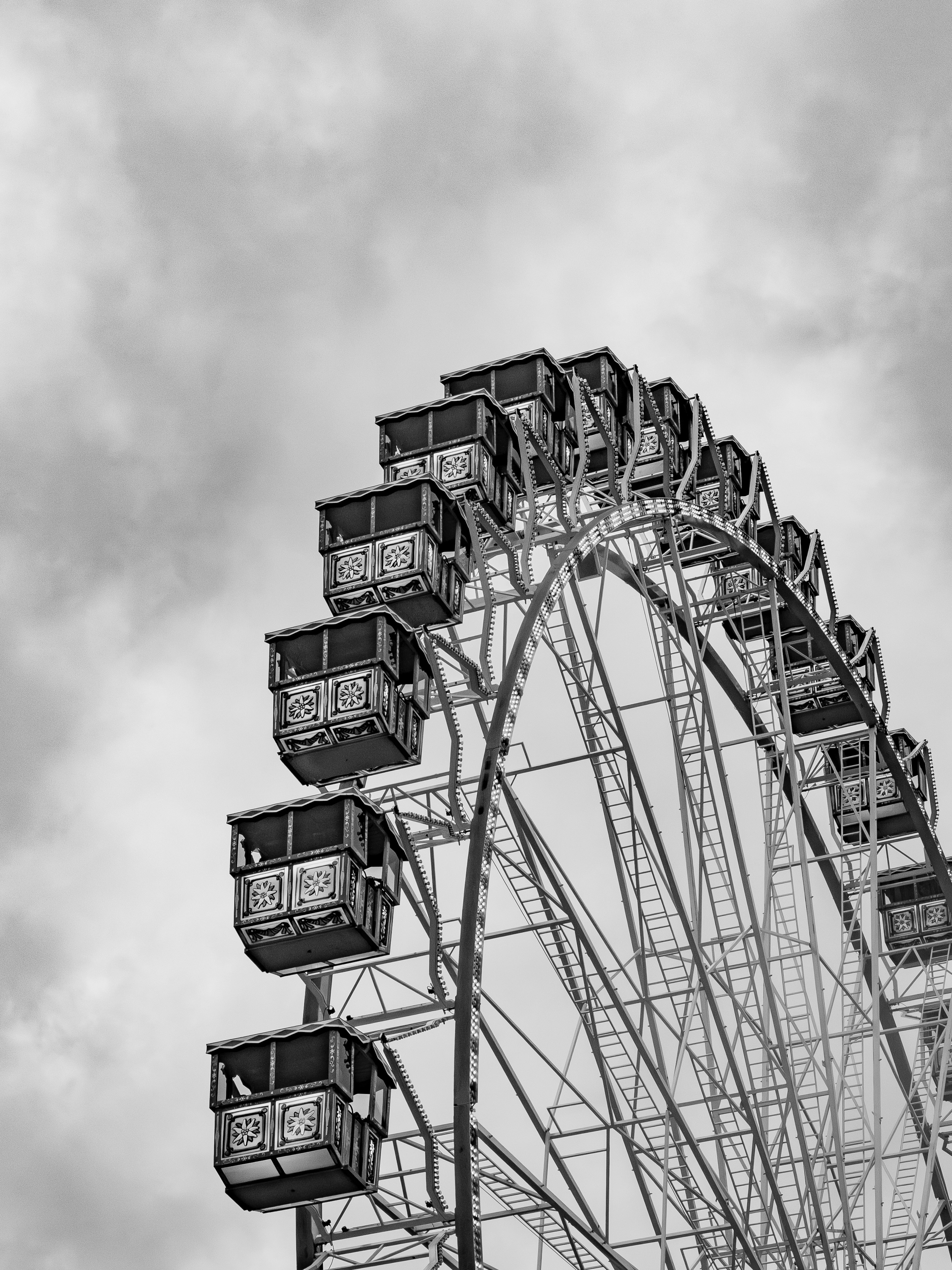 Ferris wheel rises against a cloudy sky, its intricate cabins forming a repeating lattice. A monochrome capture emphasizes structure and scale, with the wheel anchored as the focal element of the composition.