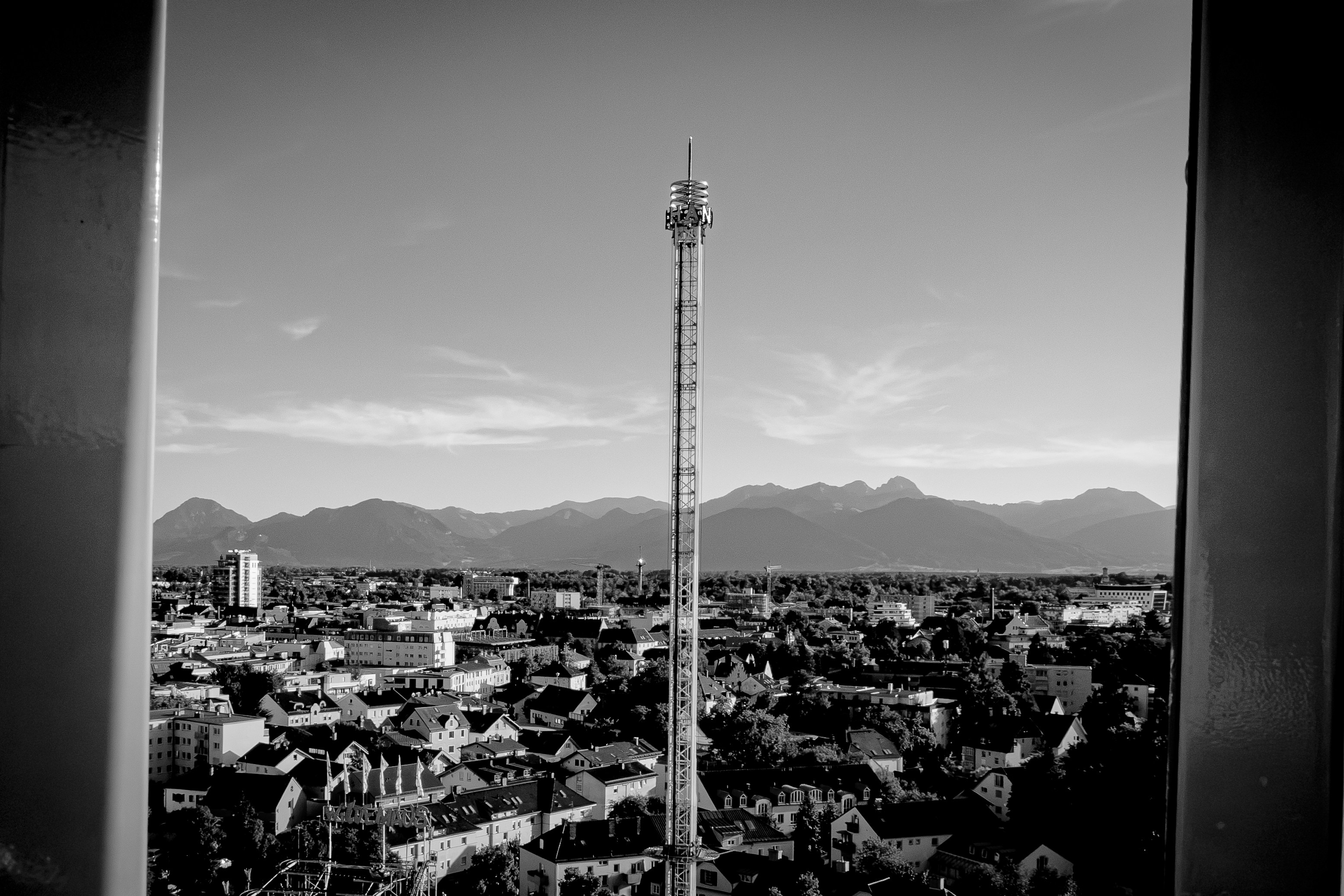 A grayscale cityscape framed by window edges, with a tall central tower rising above the rooftops, and distant mountains on the horizon.