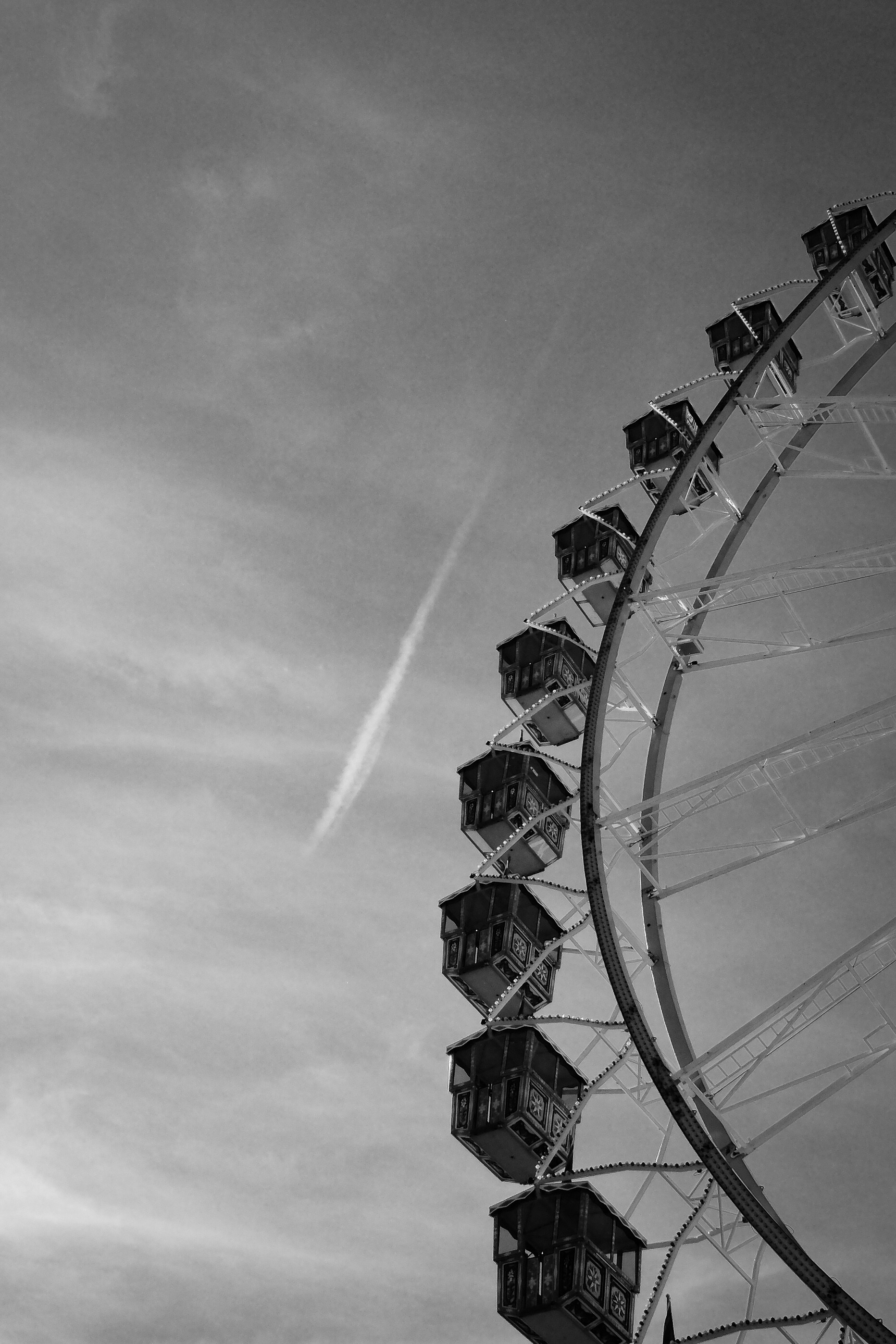 Black-and-white shot of a large Ferris wheel occupying the right half of the frame against a pale sky with a contrail crossing diagonally.