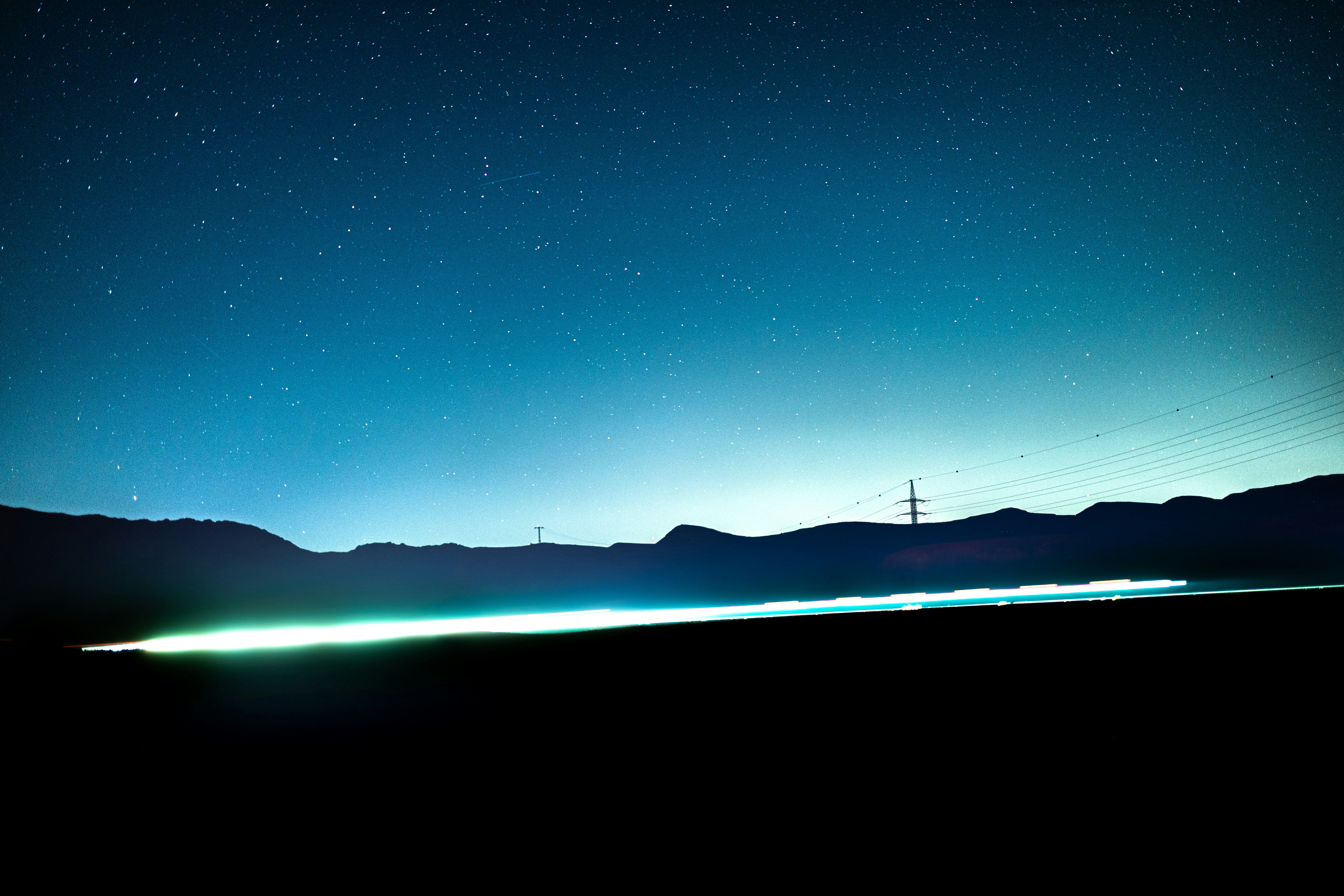 Night sky speckled with stars above a dark horizon and a glowing light trail along the ground; a minimalist landscape with distant power lines. (photography) 