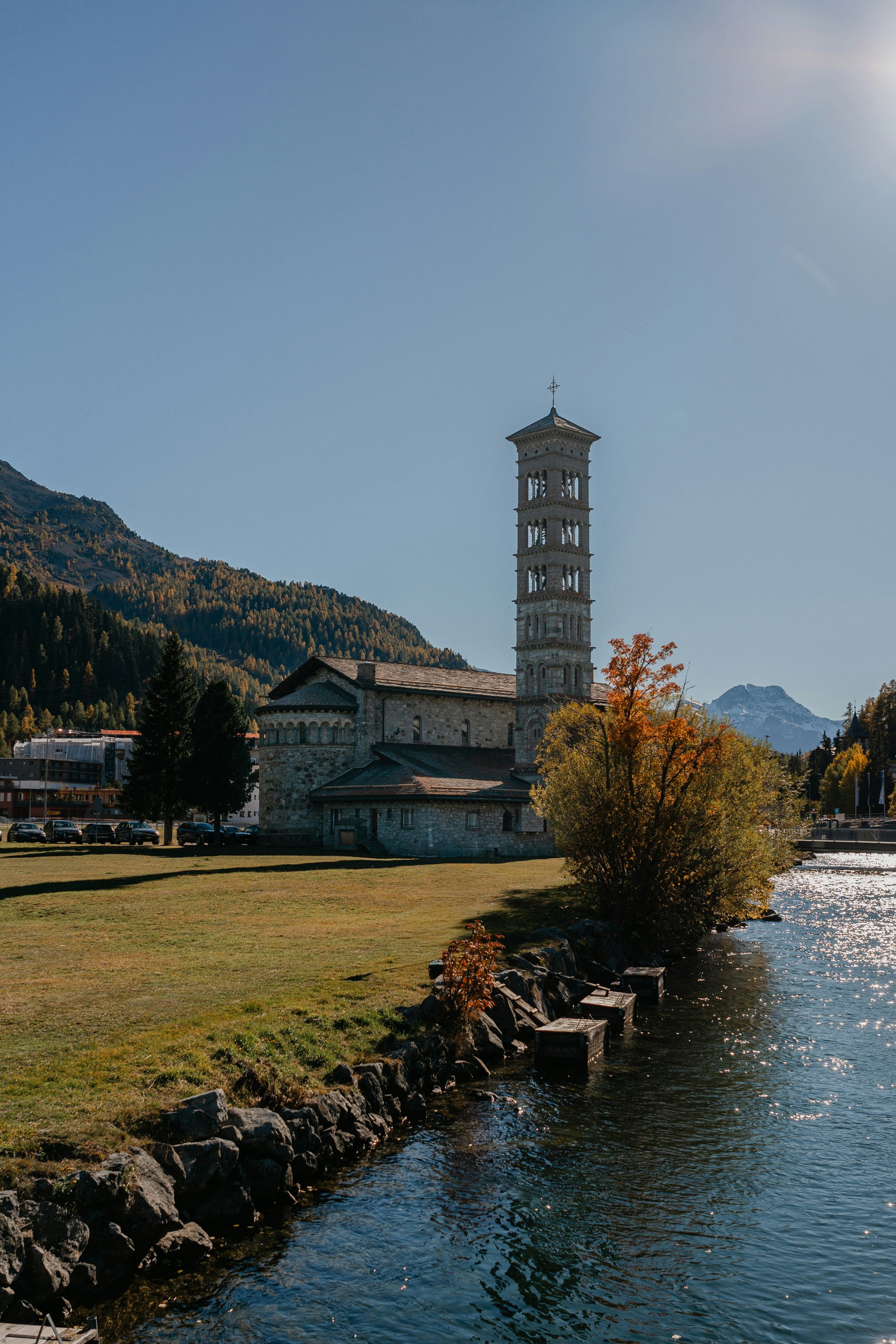 Alpine church with a tall bell tower sits beside a calm lake under clear autumn sky; warm foliage and stone walls frame the serene riverside scene.
